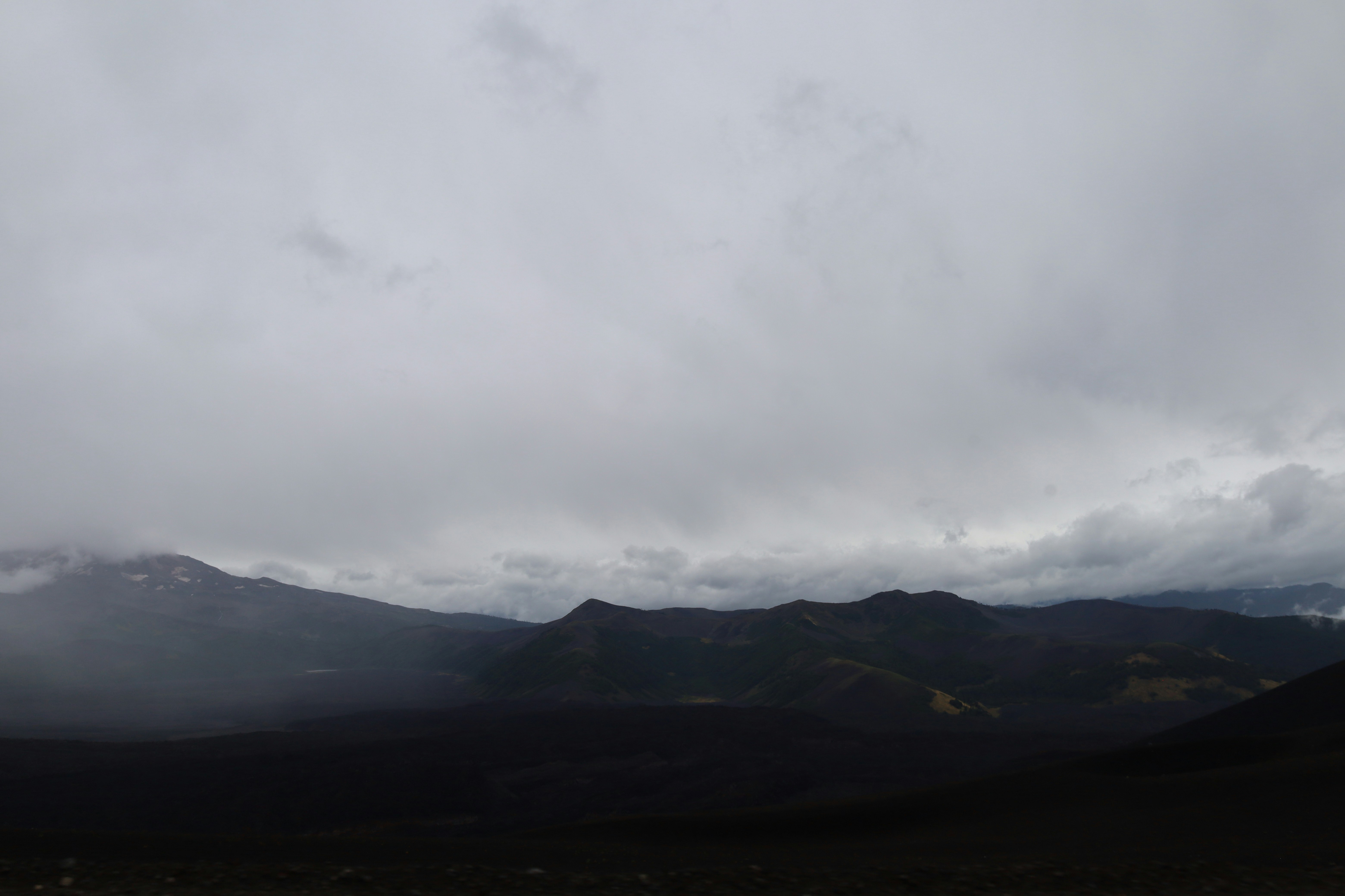 Low-contrast landscape with a dark, barren foreground and distant, cloud-cloaked ridges under a gray sky; the scene emphasizes vast atmospheric depth and subdued color.