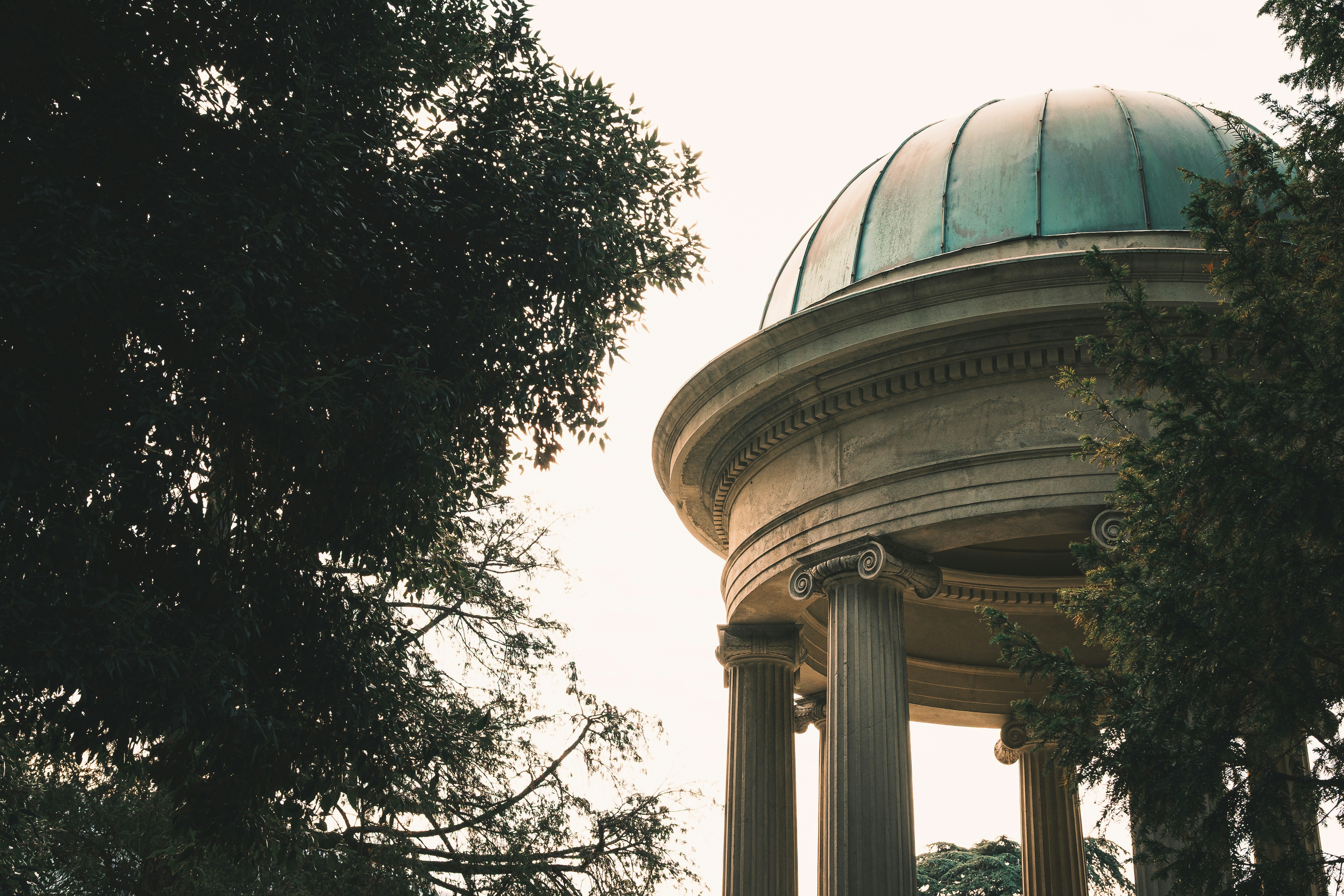 A domed rotunda with columns peeks from behind leafy trees, bathed in warm backlight, suggesting a quiet park scene. Architectural element stands as the focal feature in a natural frame.