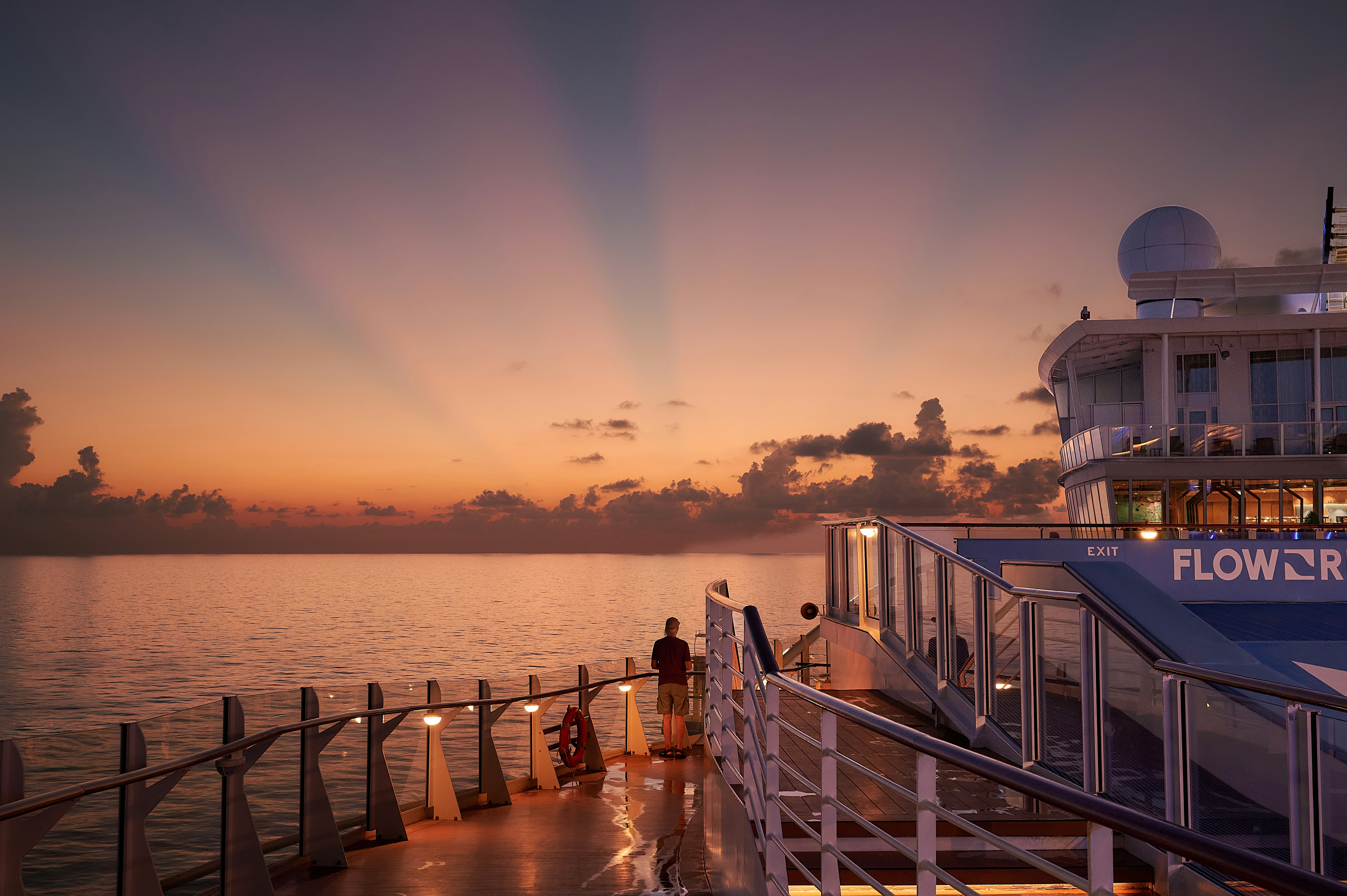 Warm sunset over a cruise deck with passengers by the railing and a calm sea to the horizon.
