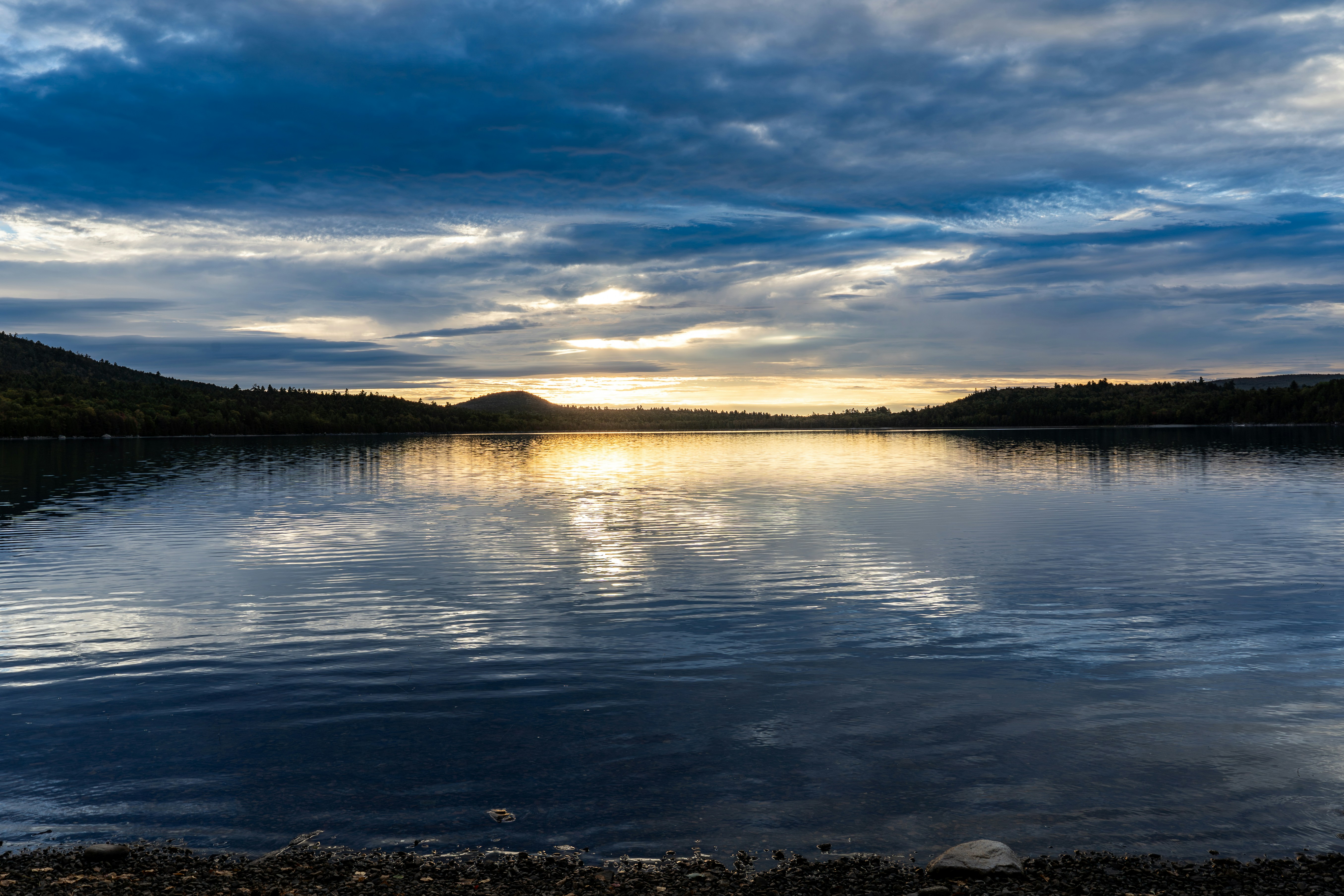 Calm lake reflecting a dramatic sky with a distant sun breaking through clouds; shoreline in the foreground and rolling hills along the horizon.