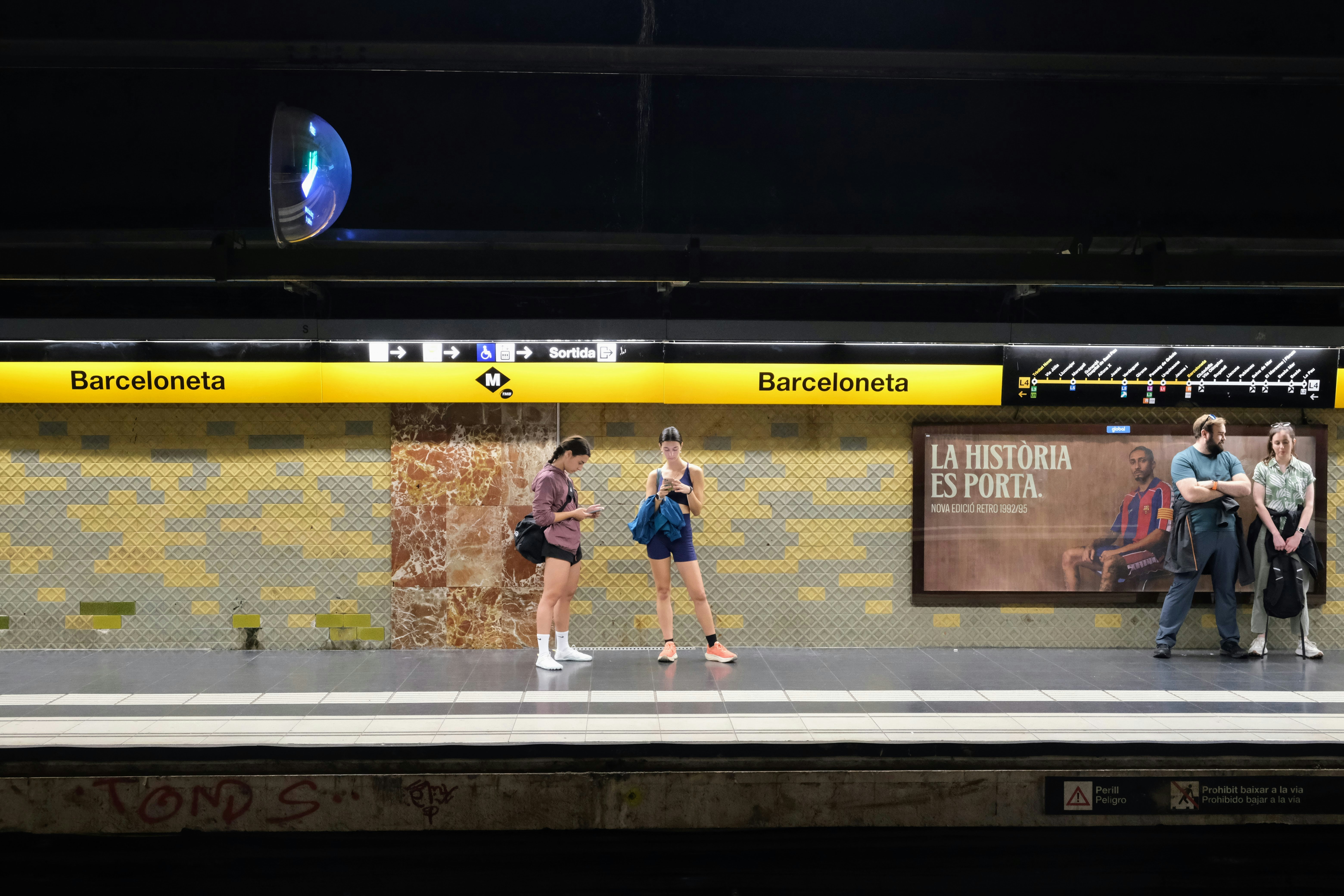 Two travelers in a sunlit metro platform pause at Barcelona equivalent signage