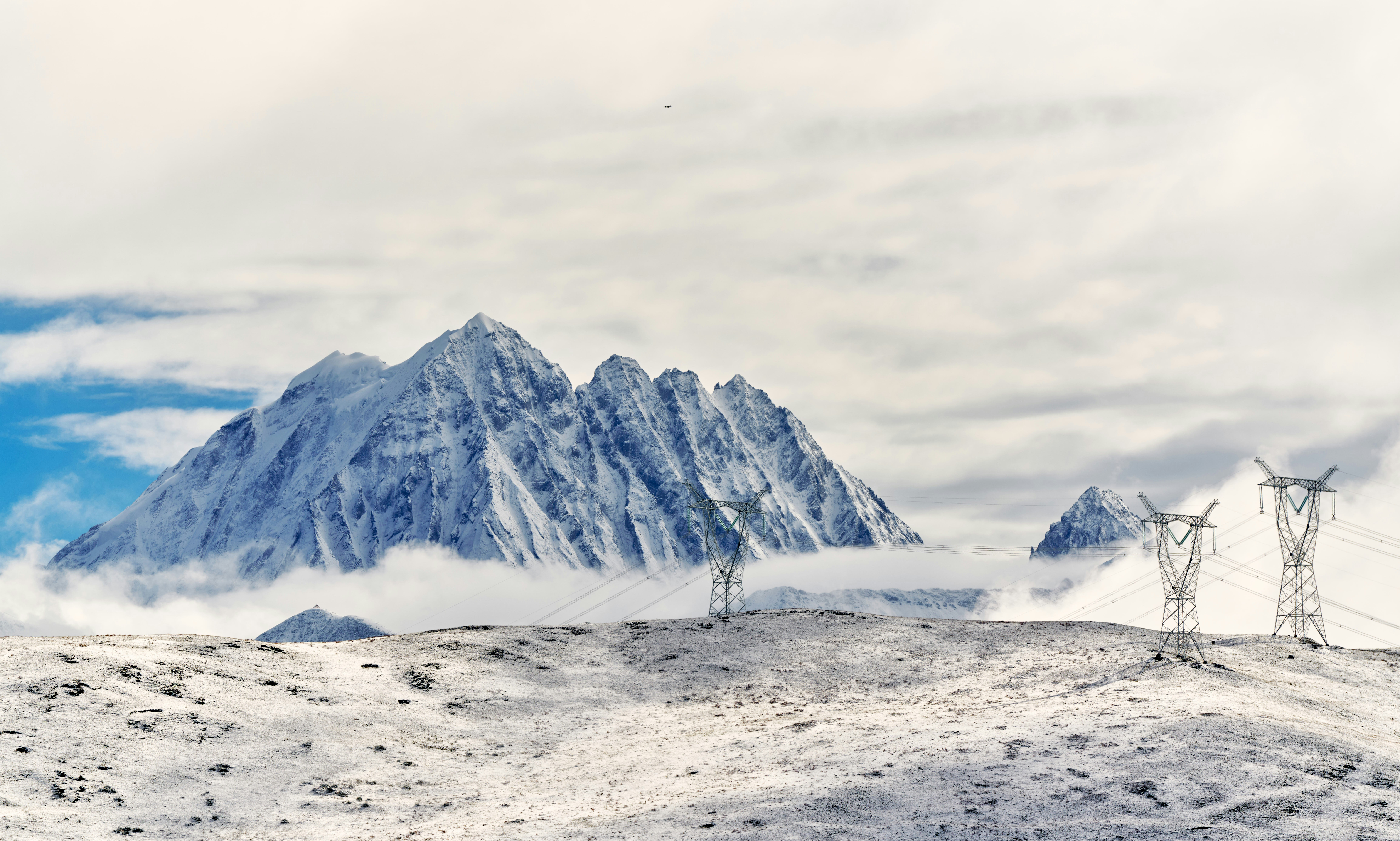 Snowy alpine landscape with jagged peaks emerging above a low cloud bank, framed by distant transmission towers on the frosted plain.