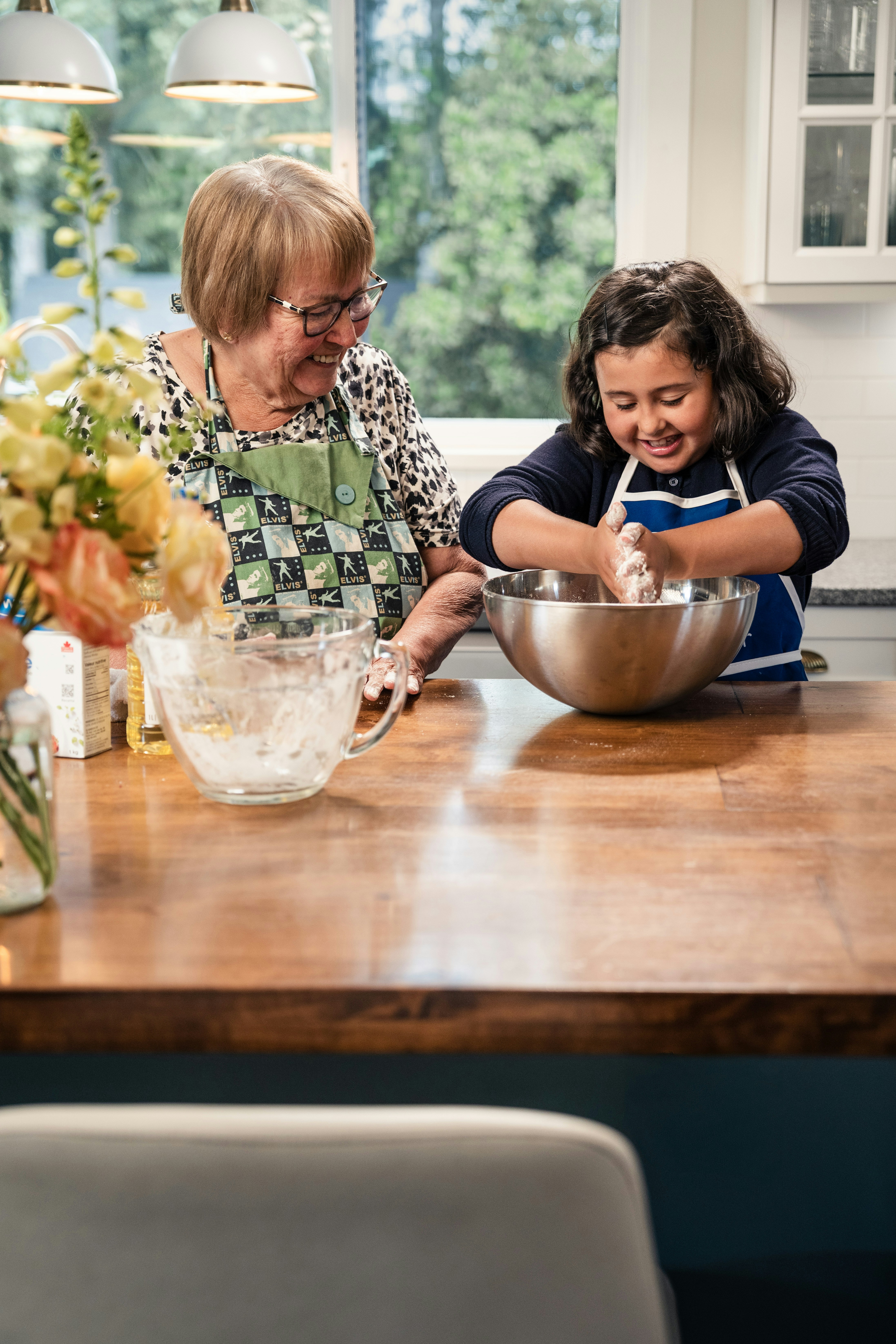 Two generations share a playful moment at the kitchen counter as flour dusts hands while mixing batter; a metal bowl and measuring jug sit nearby, framed by a sunlit window.