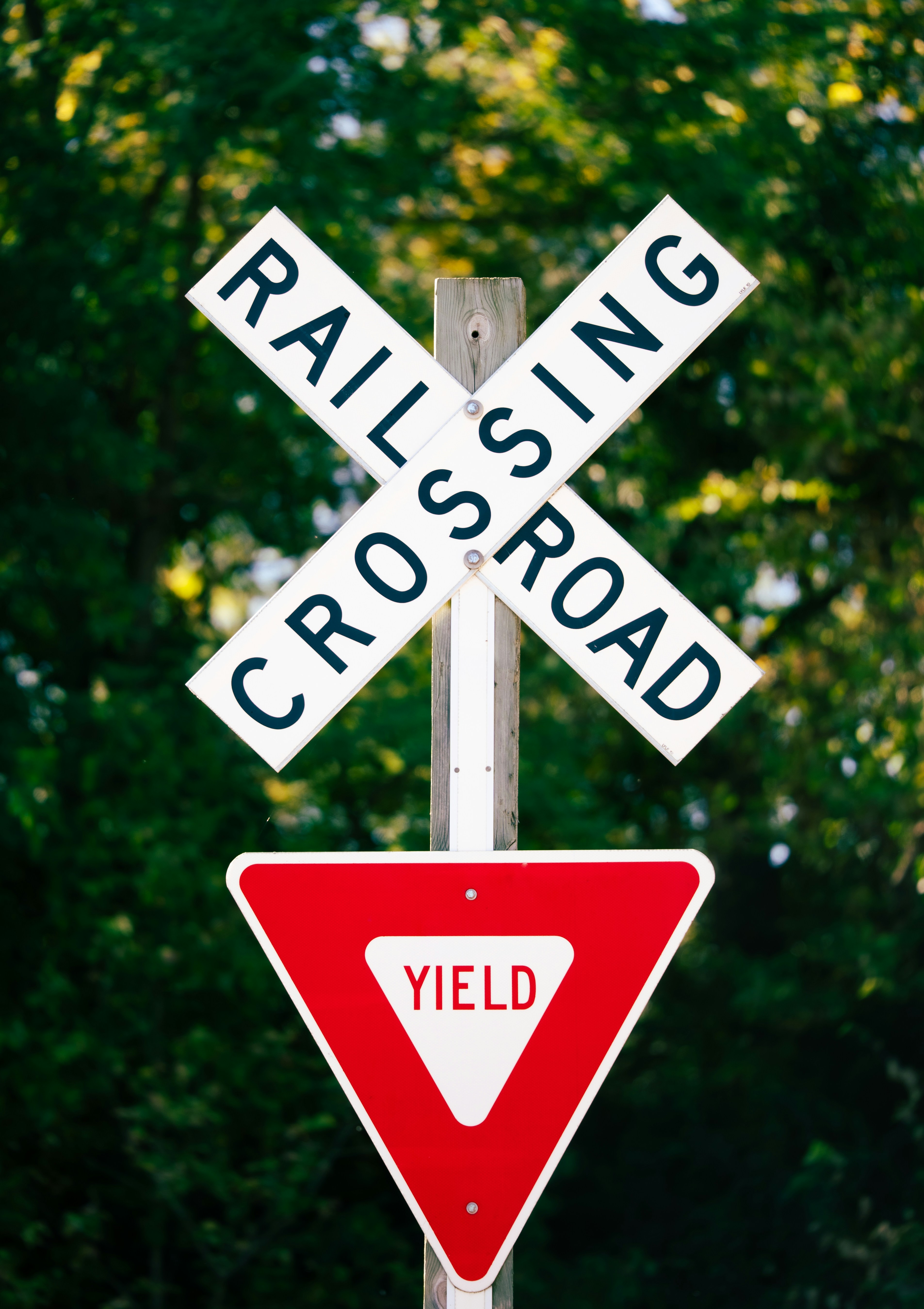 A railroad crossing sign with a red yield triangle sits against a backdrop of leafy trees, capturing a still moment of roadside warning. This is a standard street-sign composition.