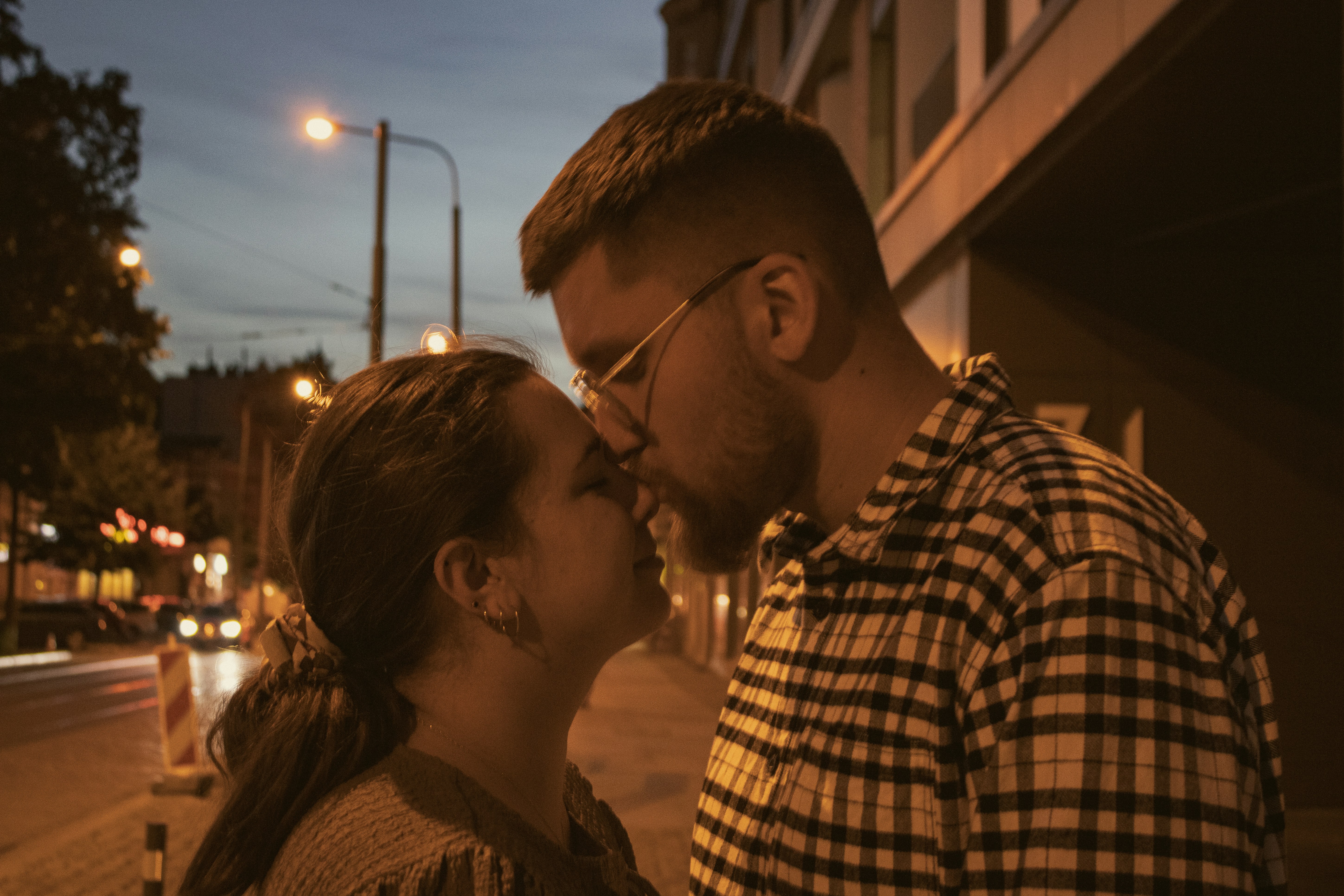 Couple leans in for a kiss on a dimly lit urban street, warm streetlamps casting a soft amber hue on their faces.