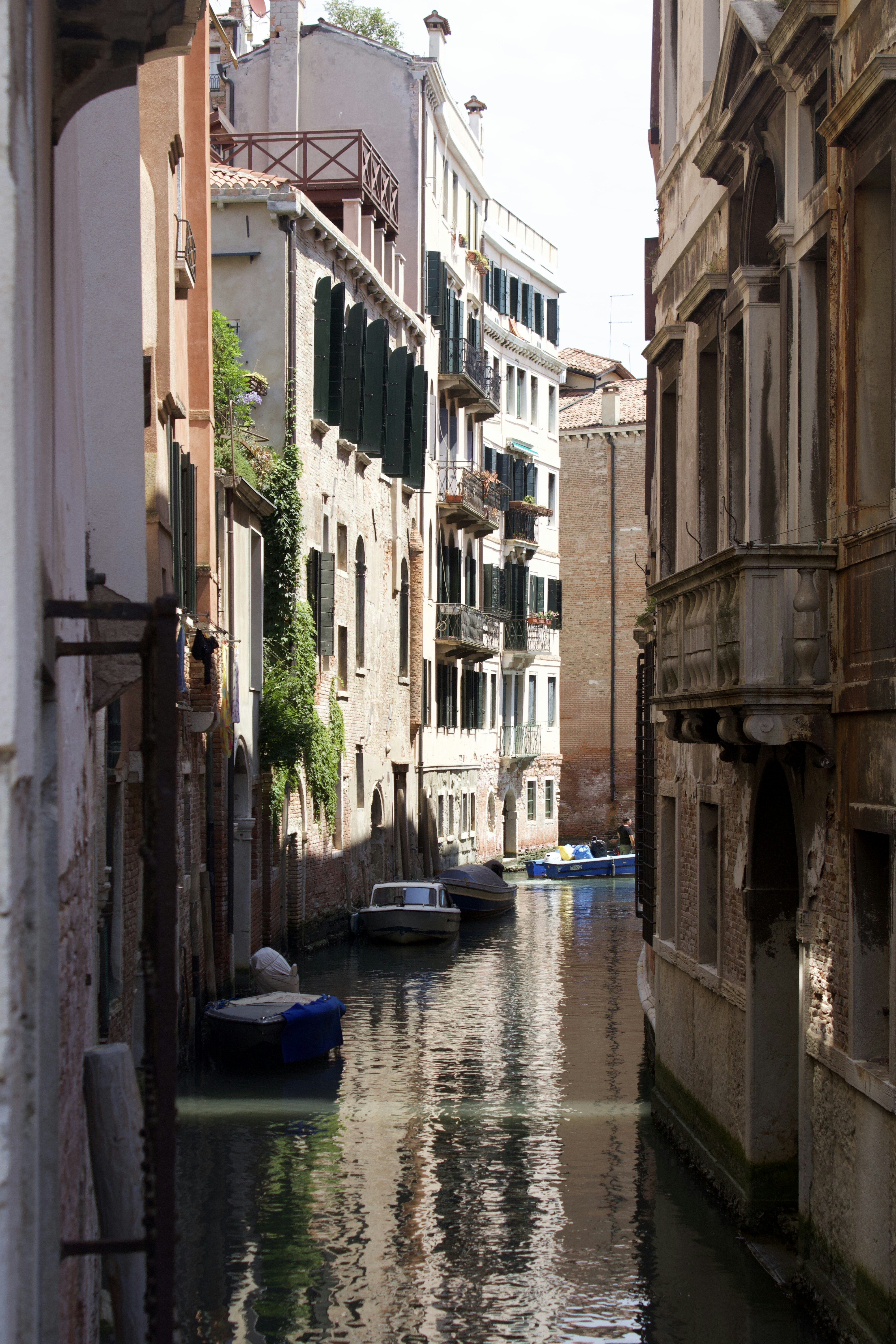 Sunlit canal flanked by aged buildings with green shutters and small boats moored along the waterway.
