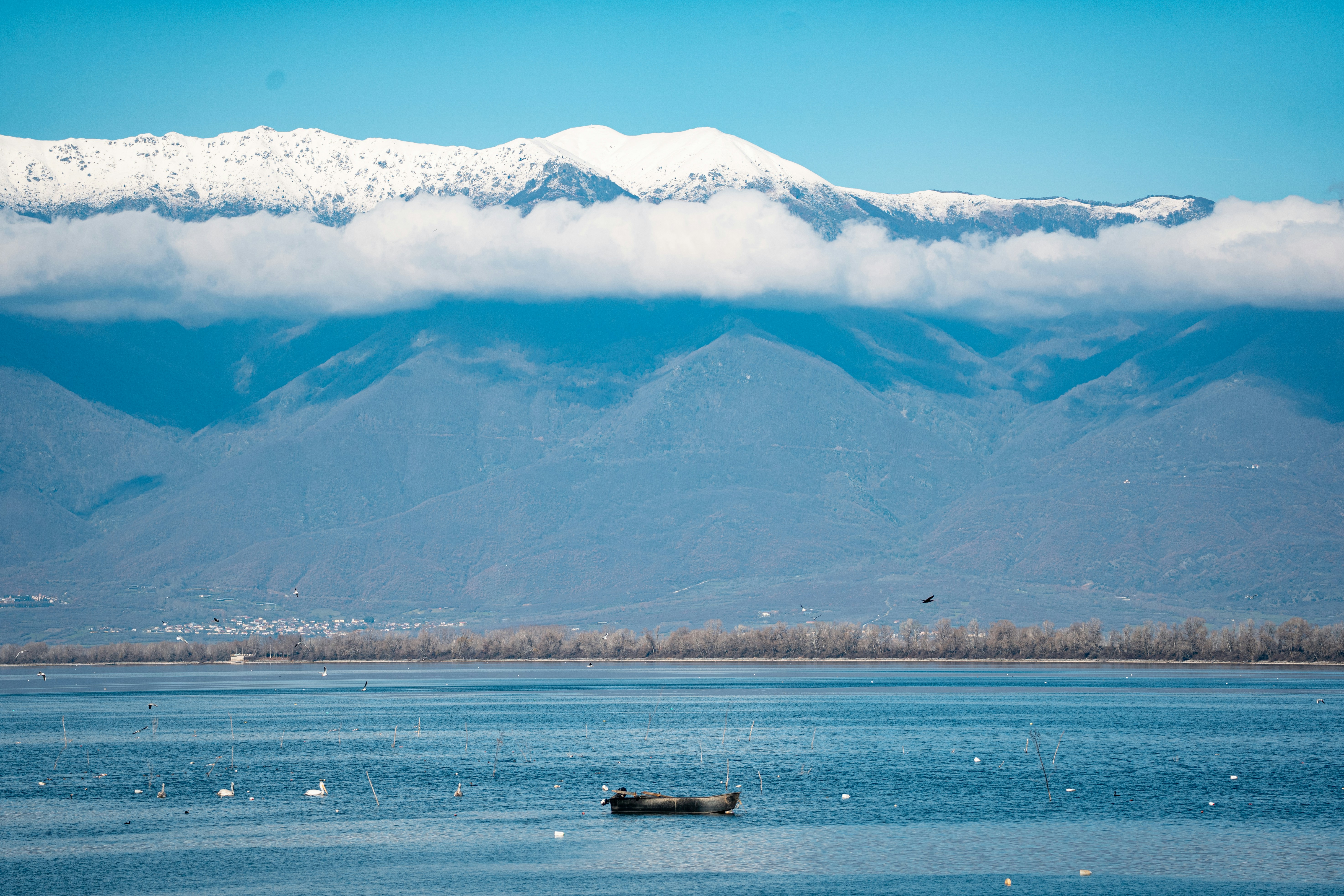 Snow-capped mountains over a calm lake with a small boat in the foreground; a thin cloud layer drifts across the mid-mountains, emphasizing depth and distance.