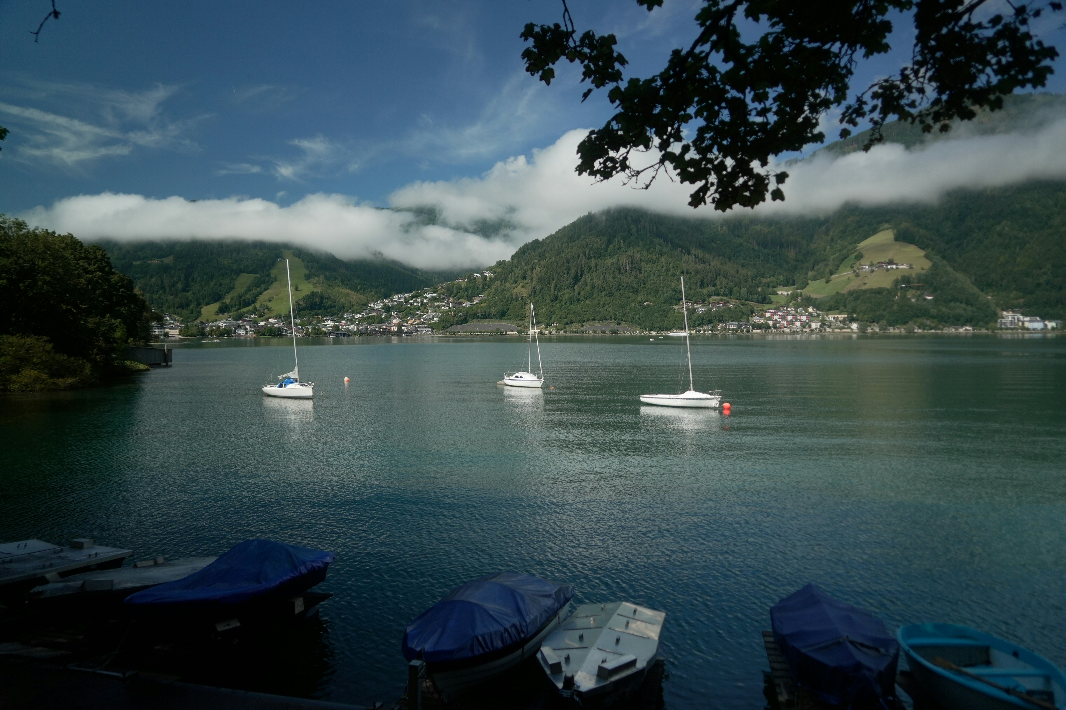 Calm lake with anchored sailboats, bordered by forested hills and a distant village, under a bright sky with scattered clouds.