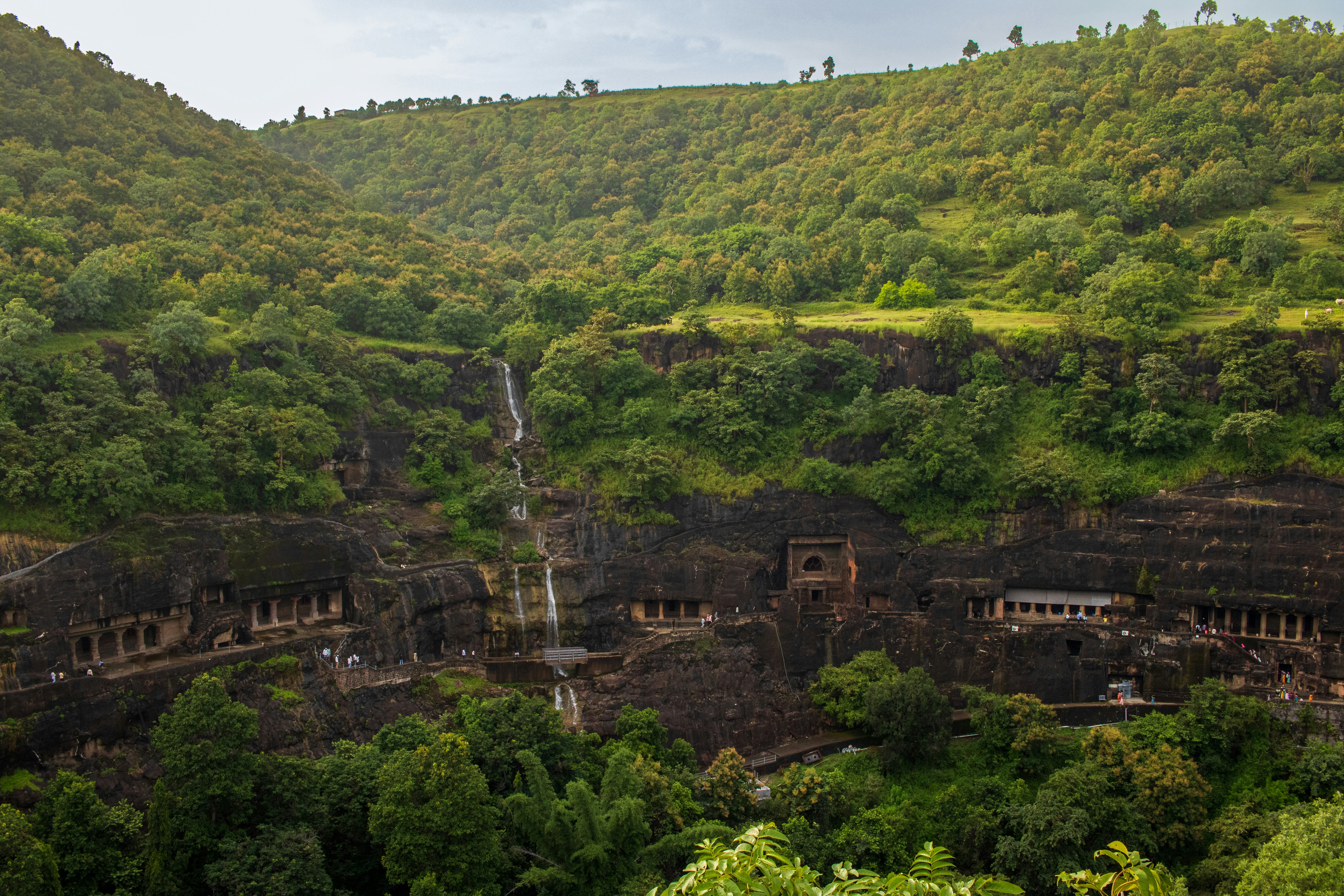 Panoramic exterior view of the Ajanta Caves carved into a horseshoe-shaped cliffside, captured from the opposite hill across the Waghora River gorge. The wide-angle shot showcases the dramatic rock-cut cave facades, ancient arched entrances, and rugged surrounding valley landscape in Maharashtra, India.