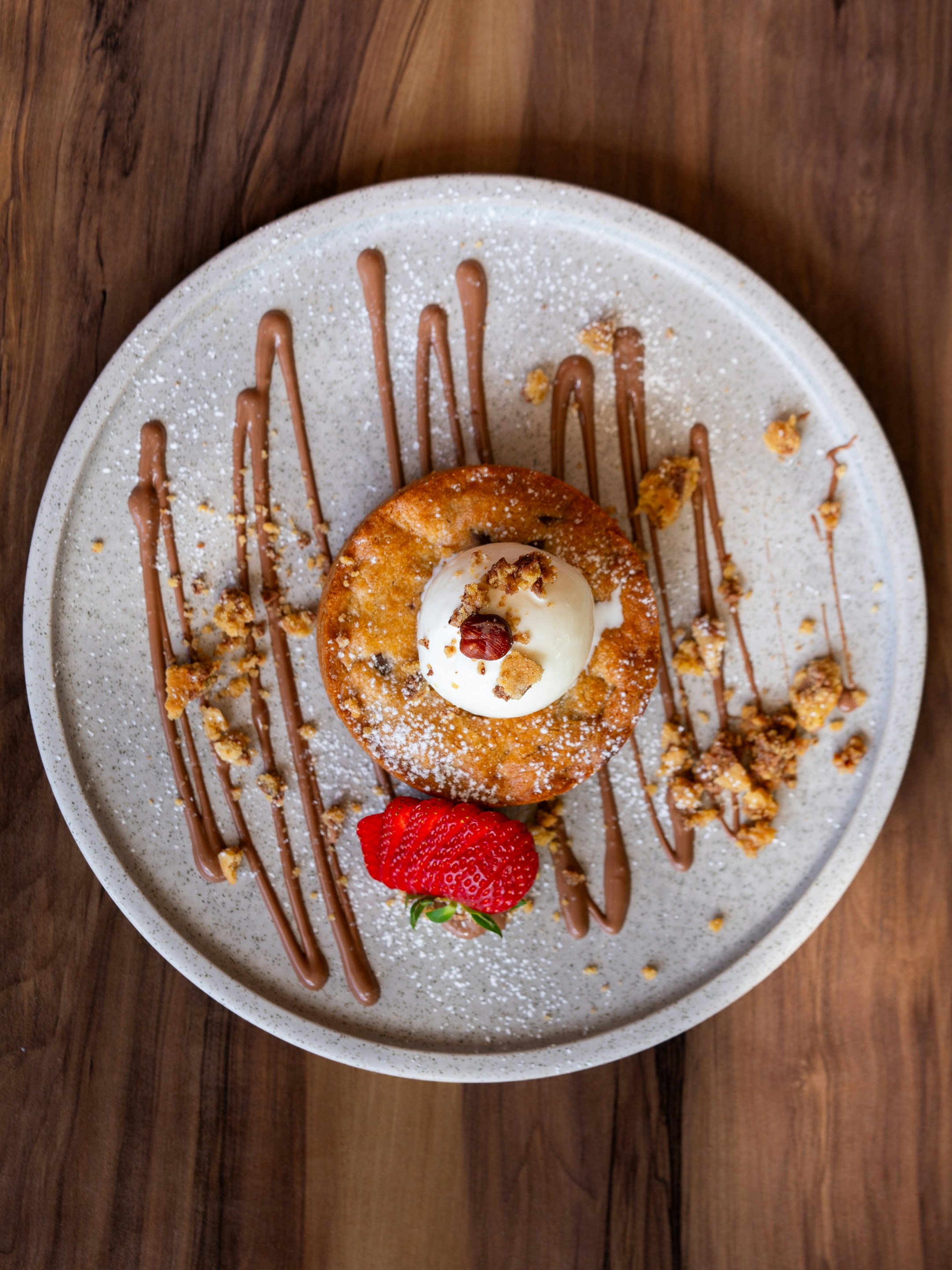 A beautifully presented sweet brunch dish served with fresh strawberry, cream, chocolate drizzle and crunchy toppings on a ceramic plate. Photographed from above on a warm timber table for a simple café-style food setting.