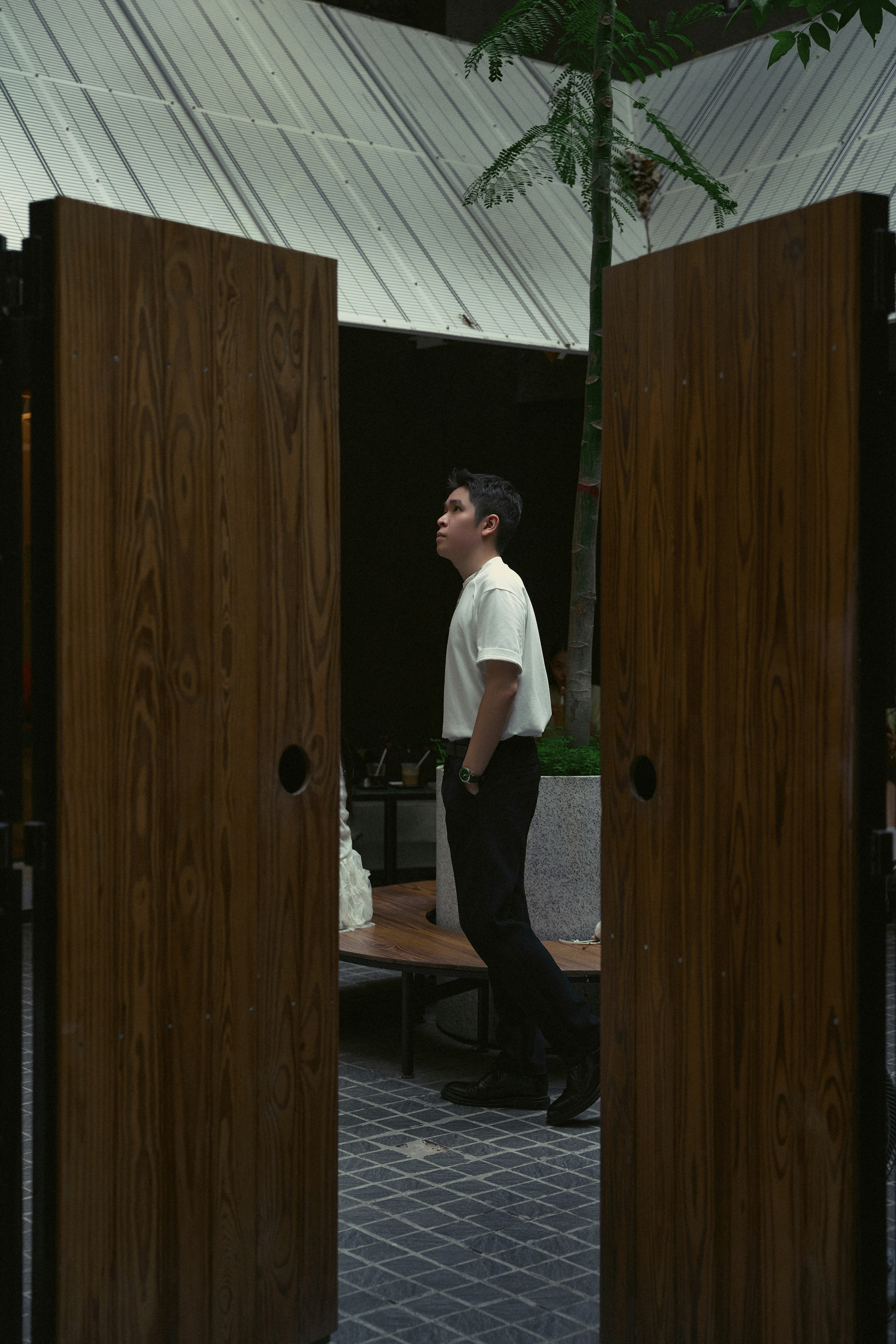 A man stands between tall wooden doors in an indoor setting, spotlighted by soft light and framed by architectural elements; the scene emphasizes calm contemplation.