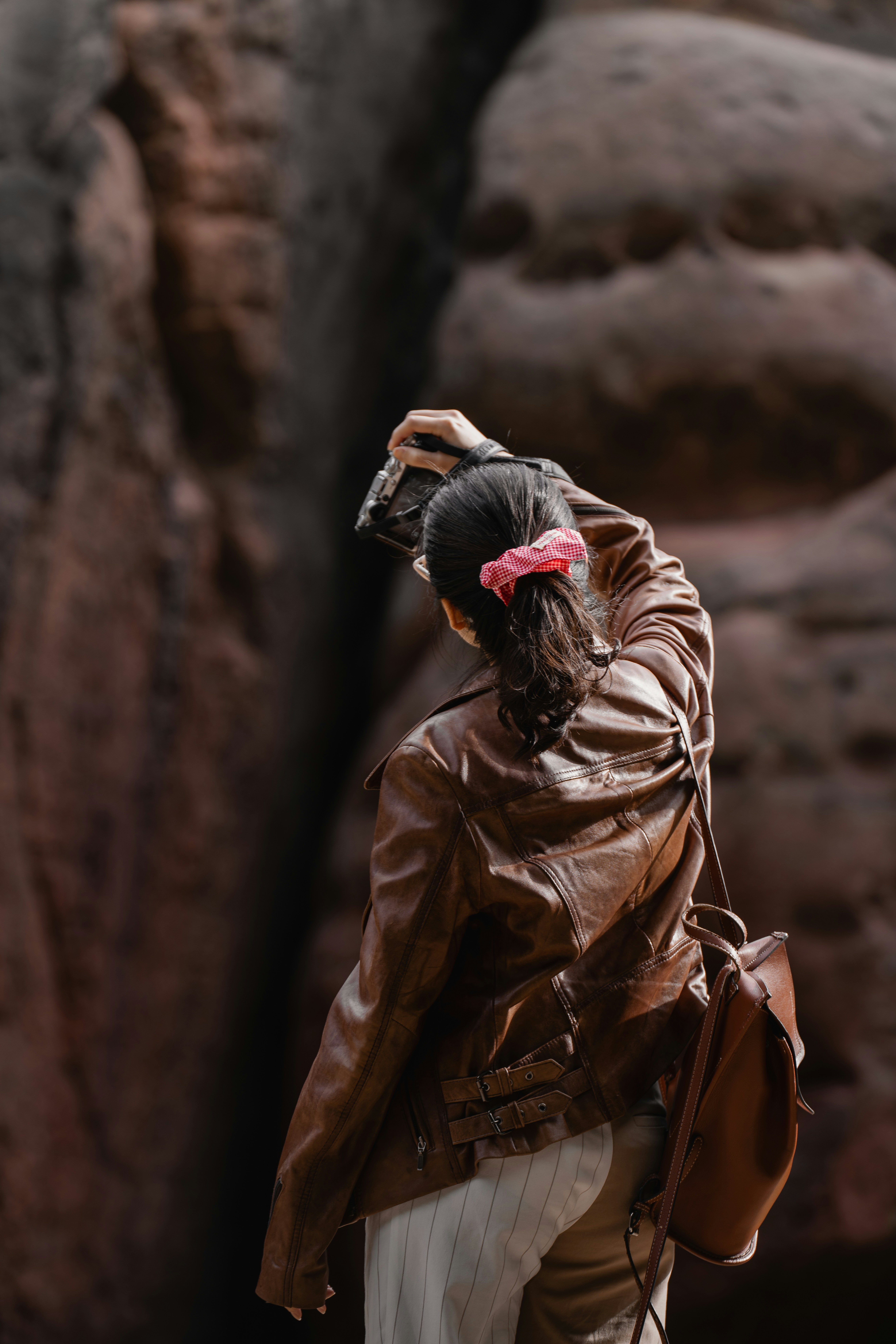 A female photographer in a brown leather jacket holds a camera up to her eye, standing against rugged canyon walls with a red scrunchie in her hair and a brown bag slung over her shoulder.