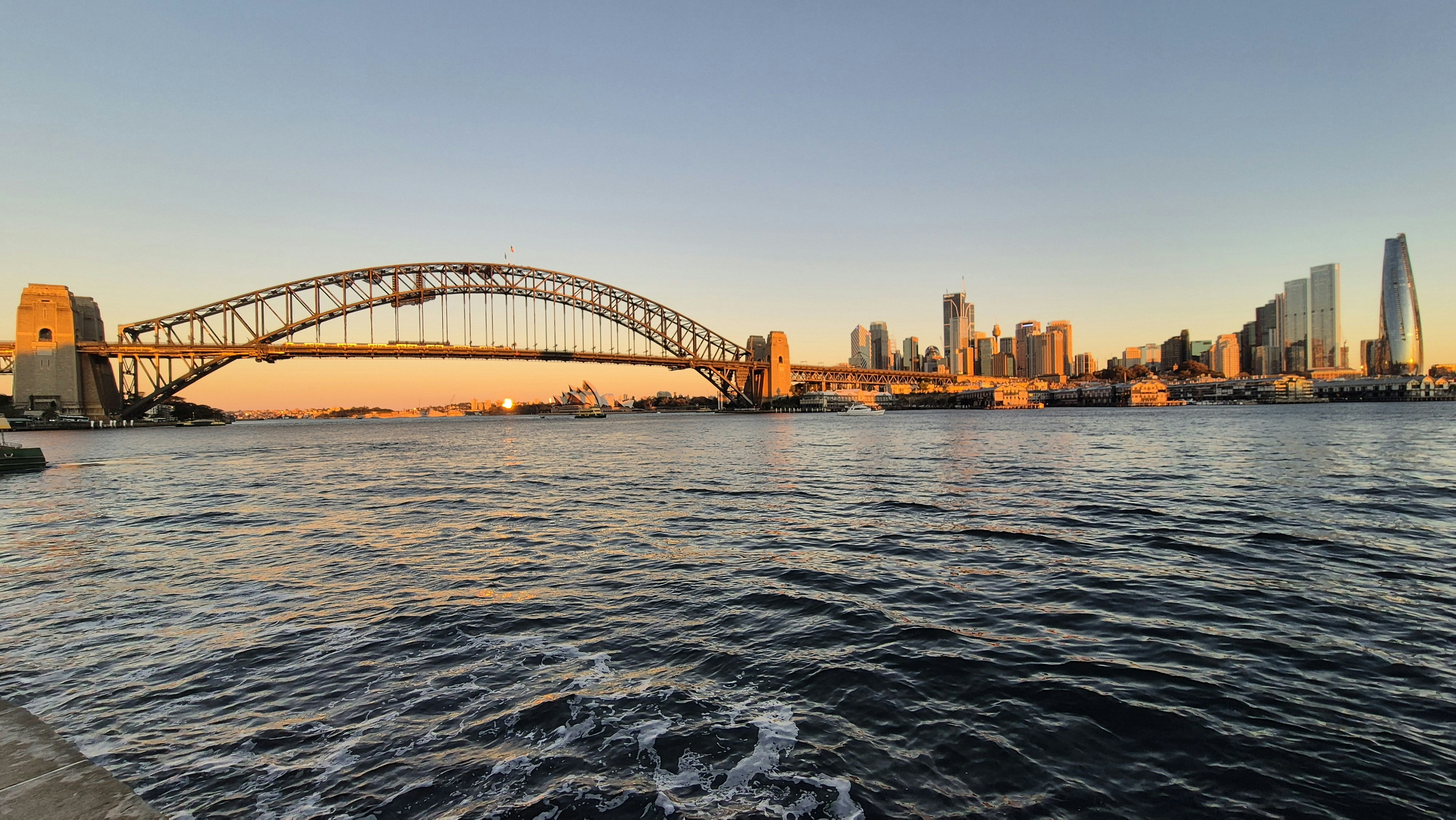 Golden hour over a harbor with a curved bridge and modern city skyline, water ripples reflecting warm light. A broad panorama featuring architectural silhouettes and a calm foreground.