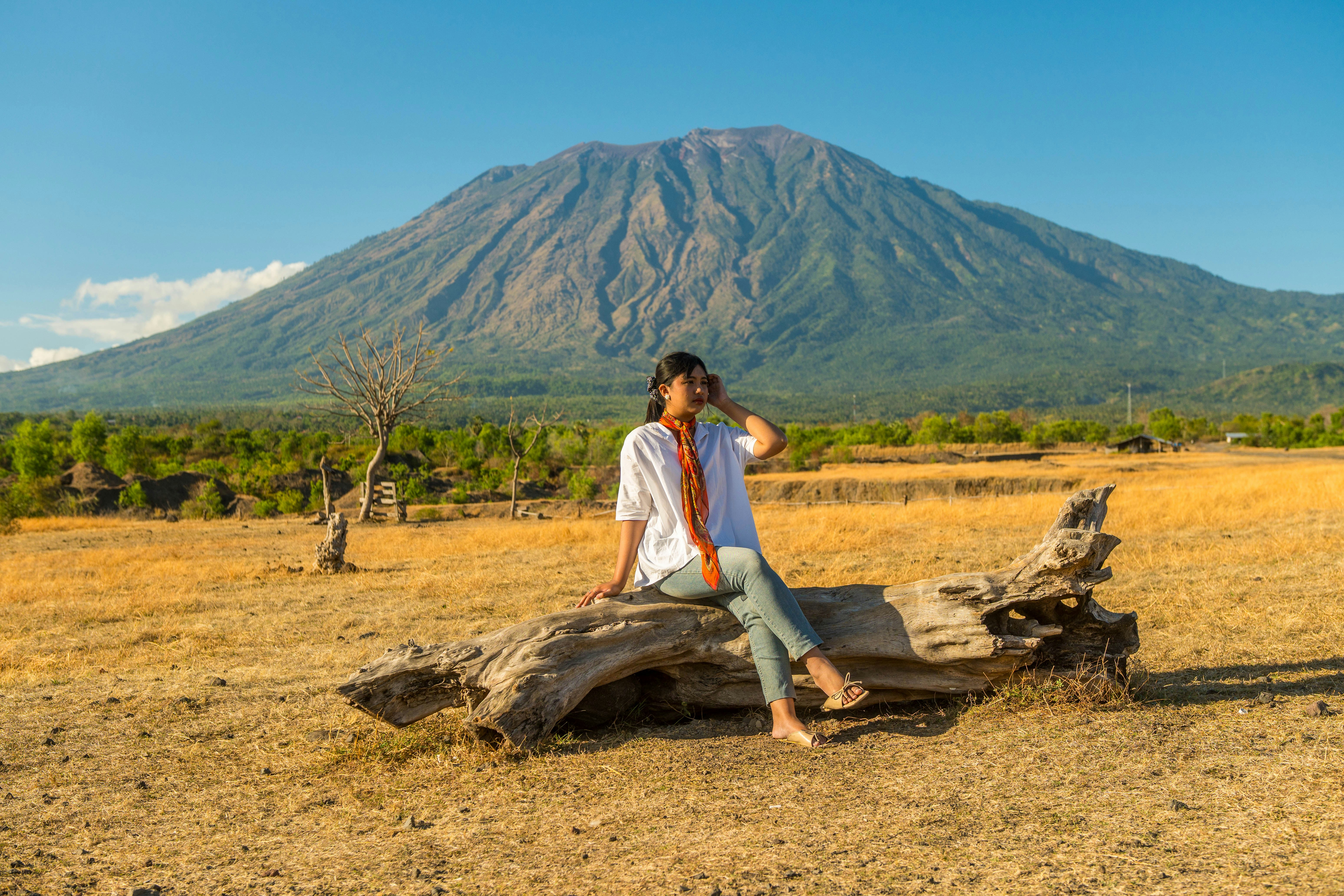 A woman sits barefoot on a weathered log in a sunlit field, with a towering volcano rising in the distance.