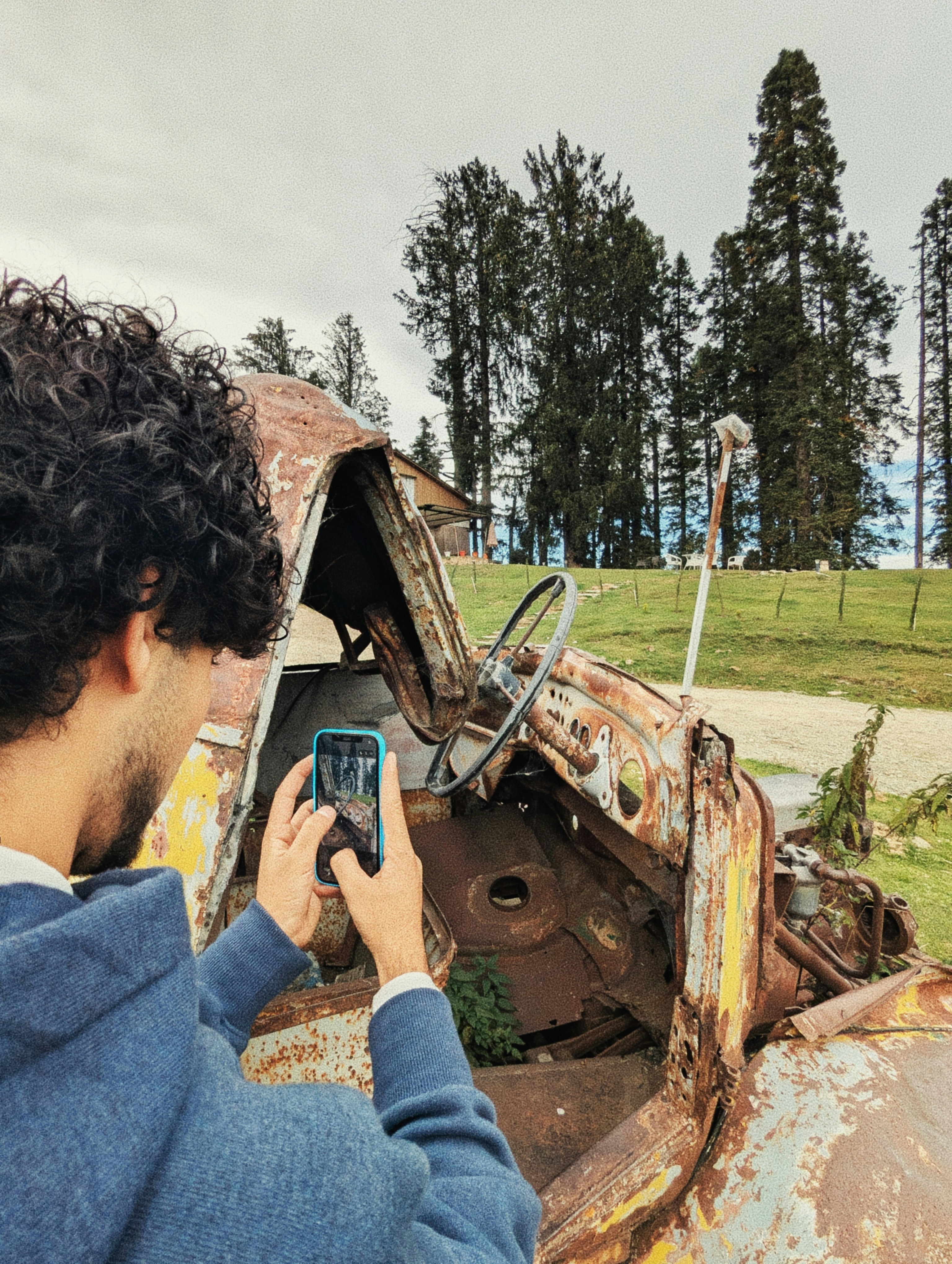 A person with curly hair holding a teal smartphone, capturing details inside a weathered, rusted vintage car parked in a grassy field with trees in the background.