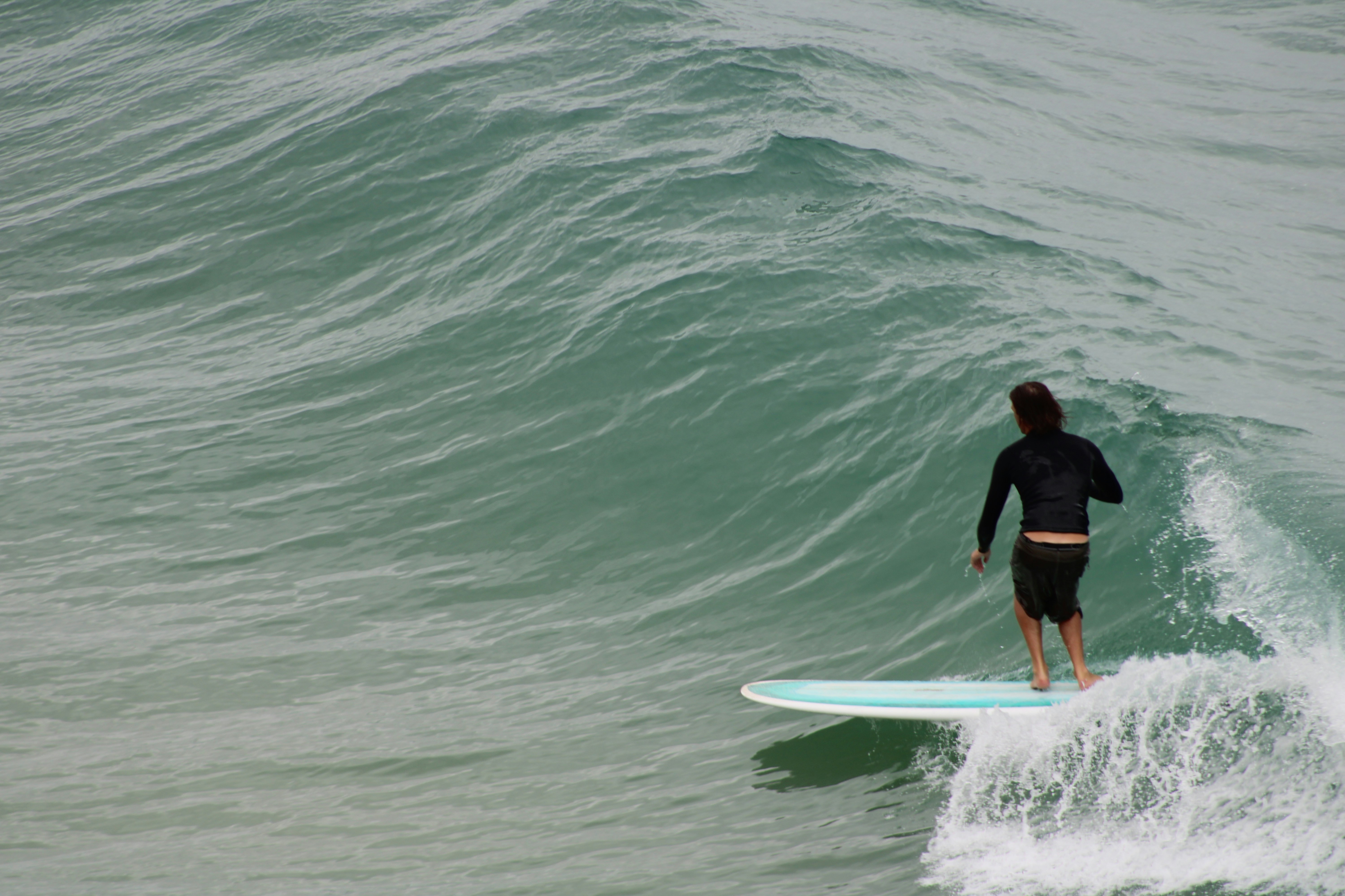 Surfer rides a clean green wave on a longboard, late in the wave with the rider positioned toward the right side of the frame.