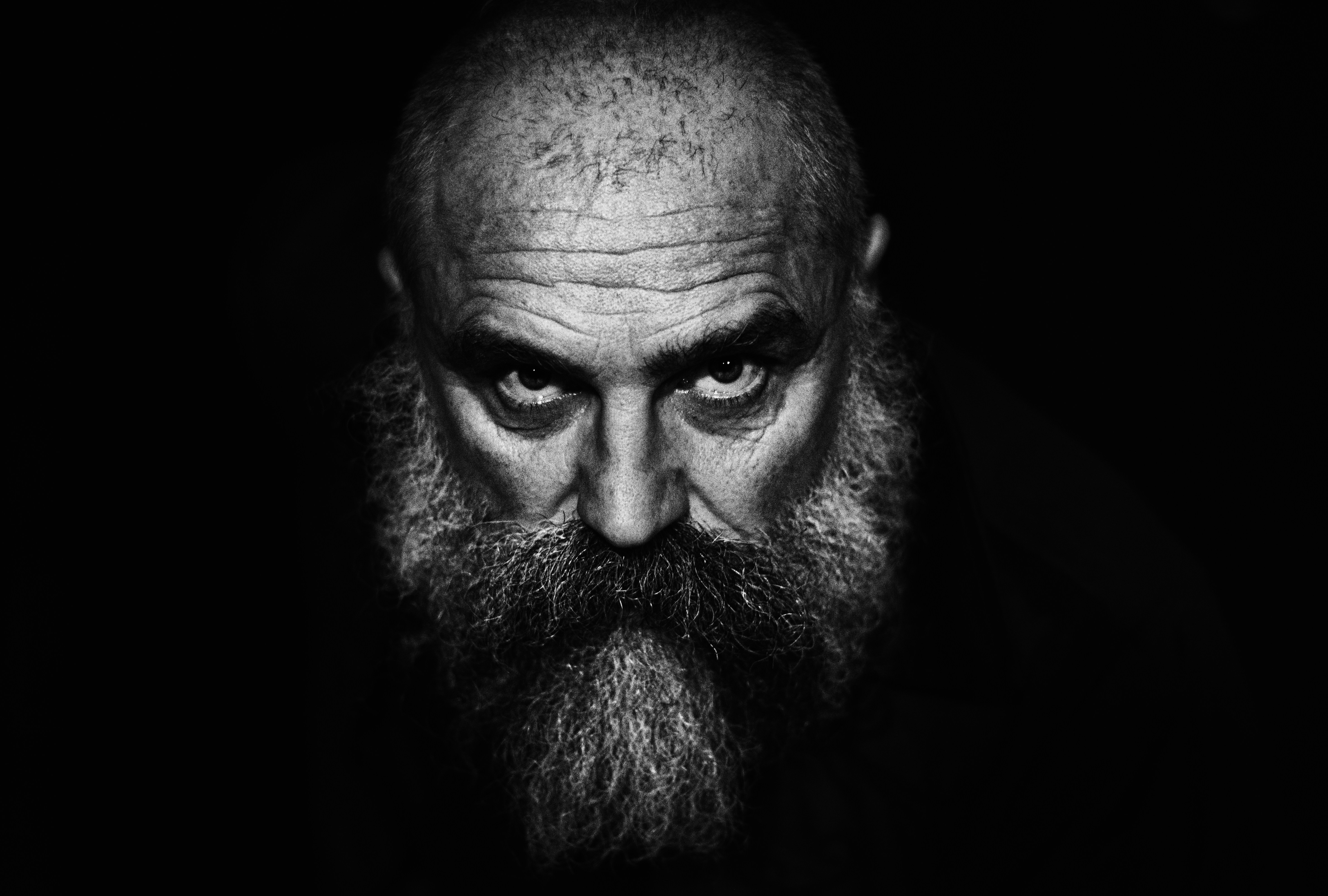 Low-key portrait emphasizing the subject's intense eyes and textured beard. The image relies on strong contrast to reveal facial features in a dark setting.