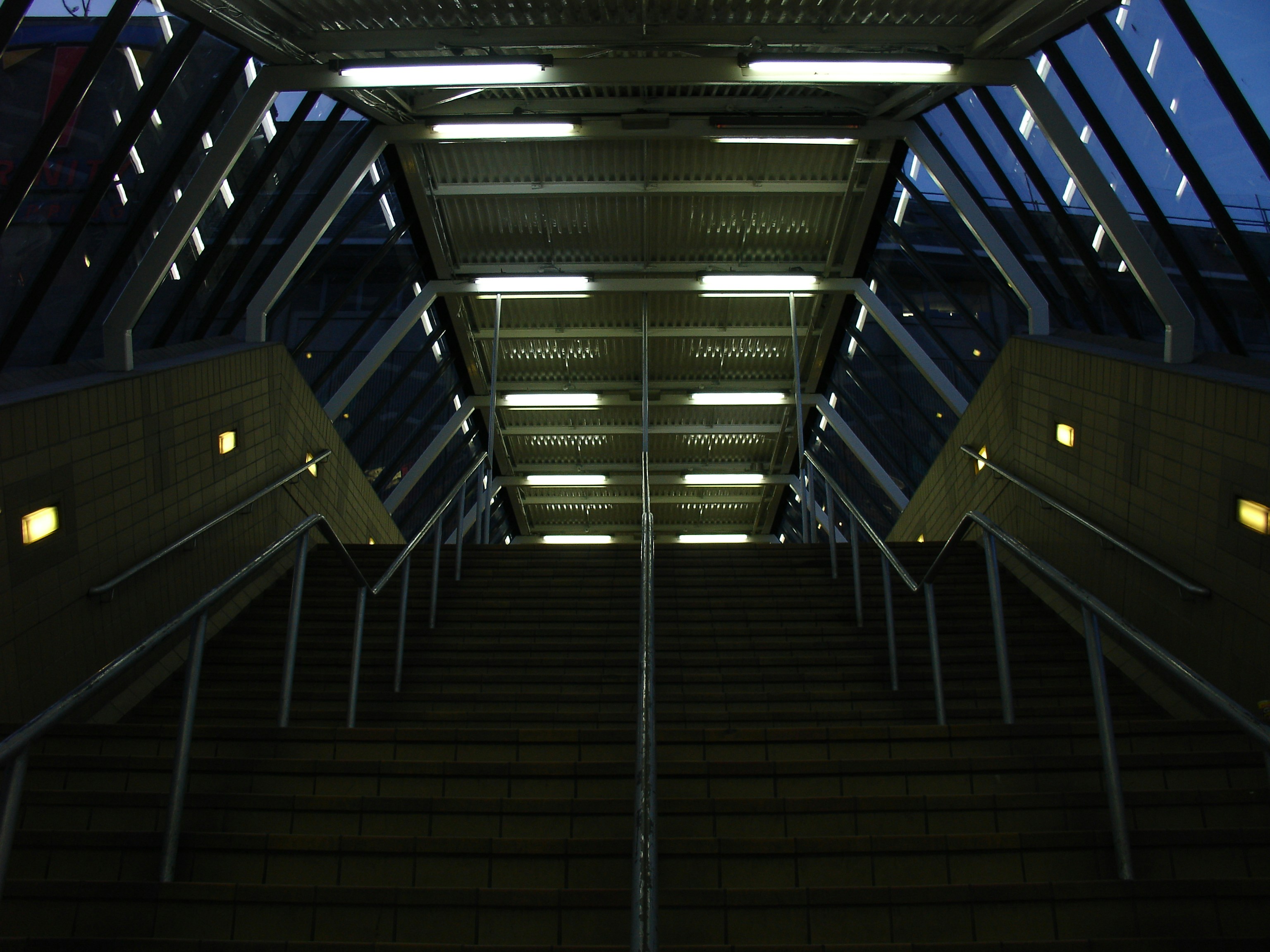 Concrete stairwell with metal railings leads upward toward a glass-roofed atrium; fluorescent strips illuminate the central path, creating a geometric, cinematic ascent.