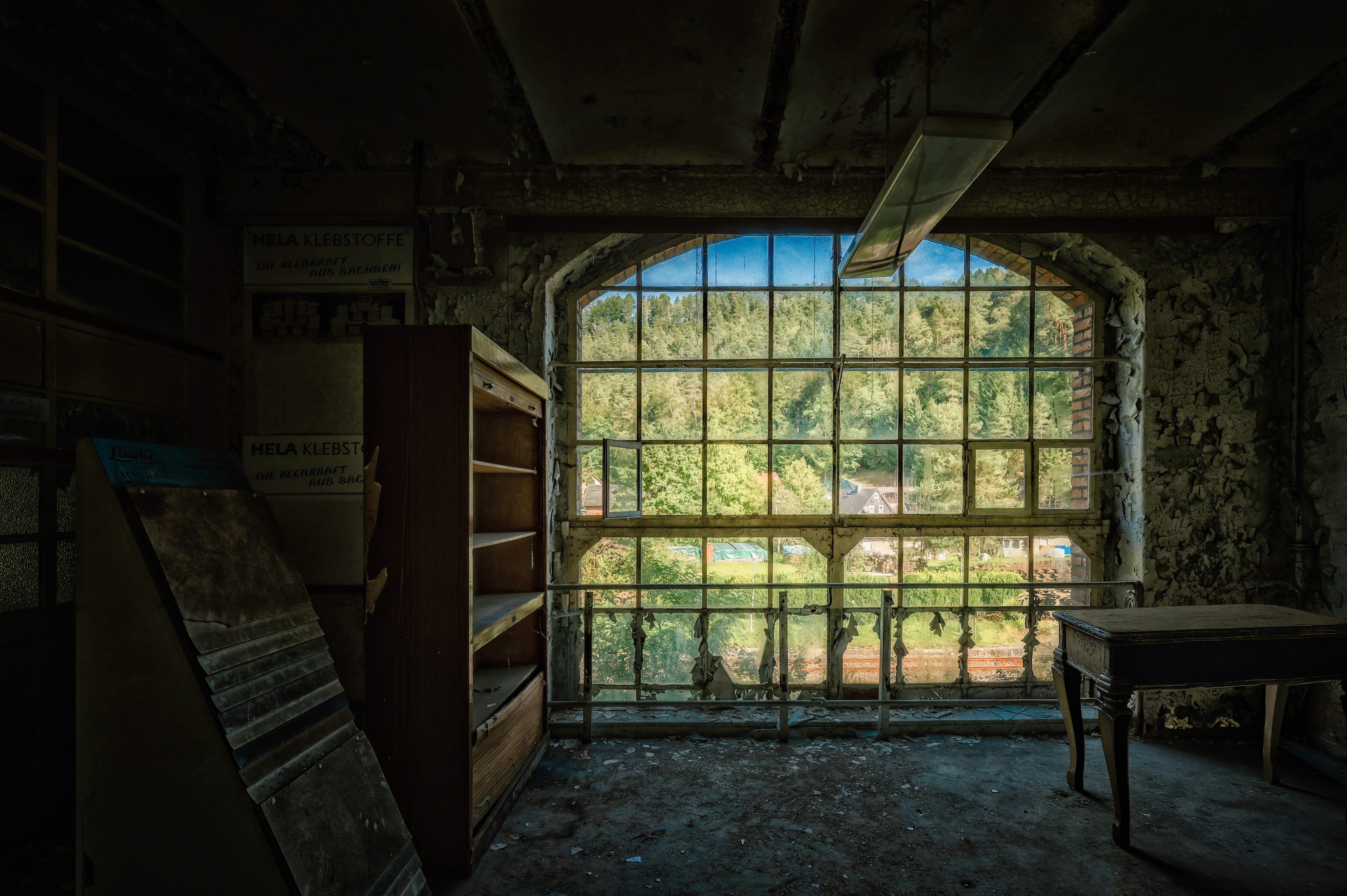 Abandoned workshop with a large arched window flooding the room with natural light; shelves, a table, and peeling walls reveal decades of neglect in a muted, moody scene.