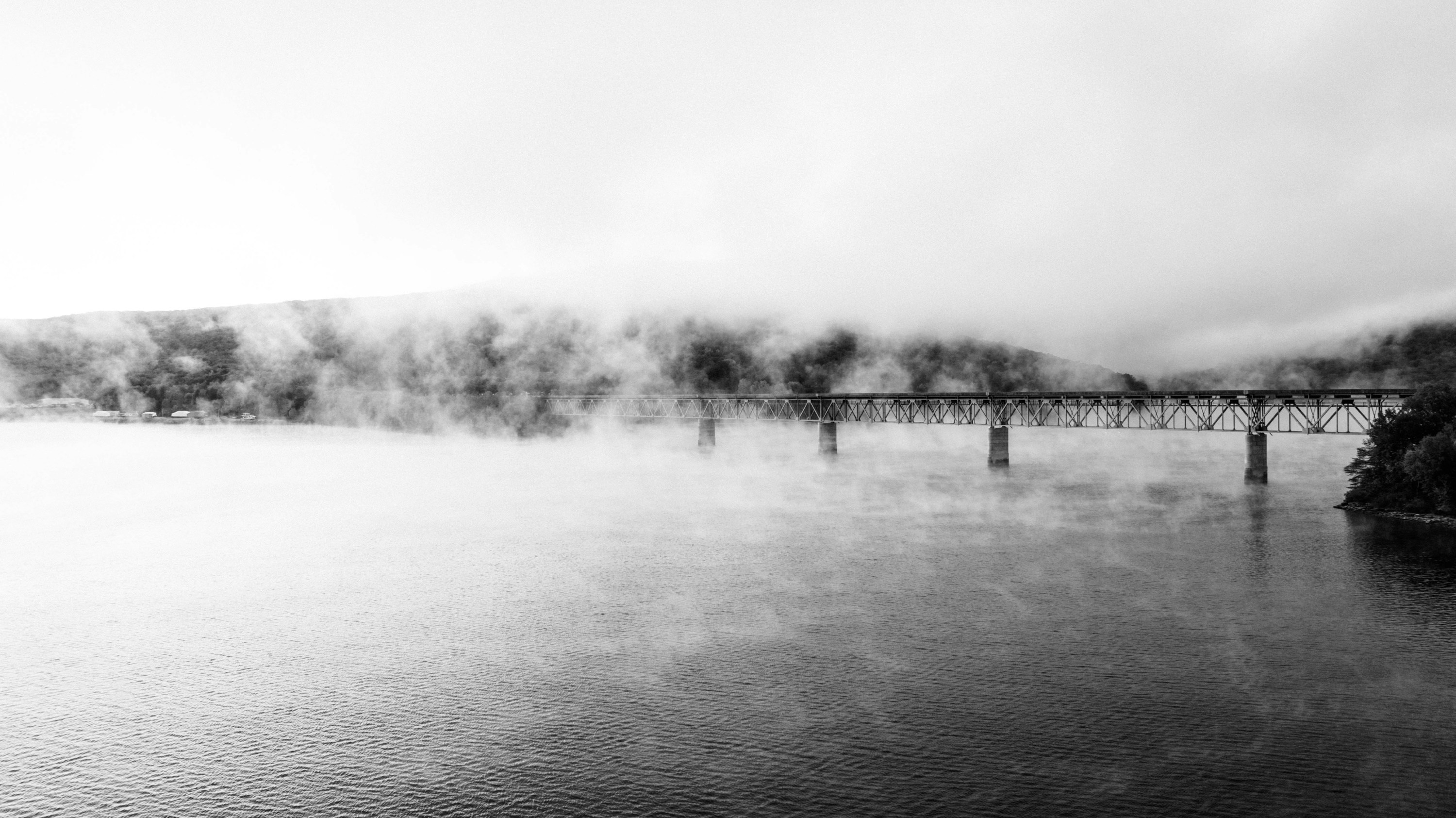 Mist clings to a tranquil river with a distant bridge, rendered in monochrome to emphasize texture and mood.