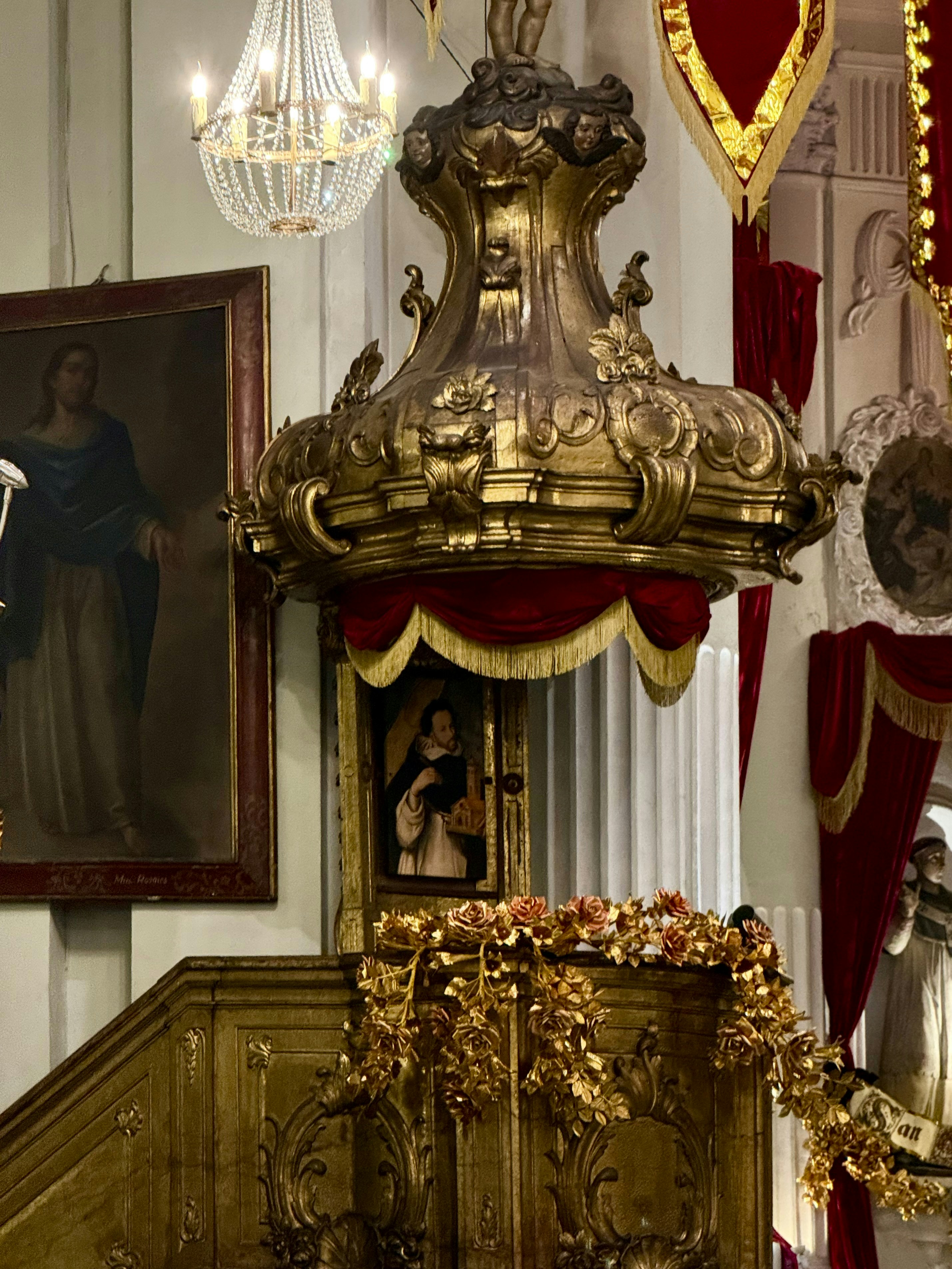 Ornate gilded altar crown with scrolling details and a small religious tableaux sits within a richly decorated interior featuring a chandelier and framed icons.