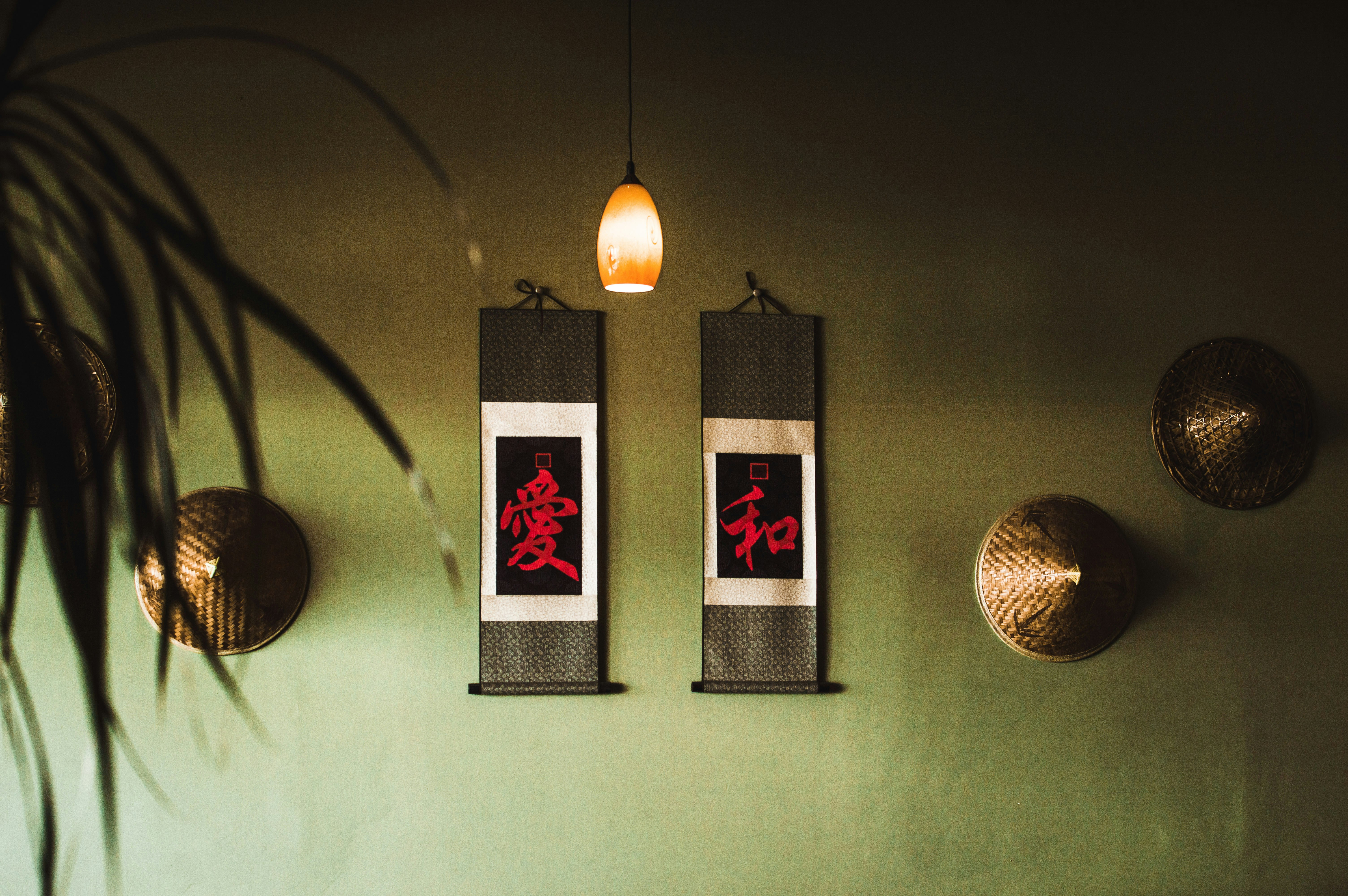 Two vertical hanging scrolls with red characters hang on a green wall, softly lit by an amber pendant lamp above; a foreground plant shadow adds depth to the intimate interior.