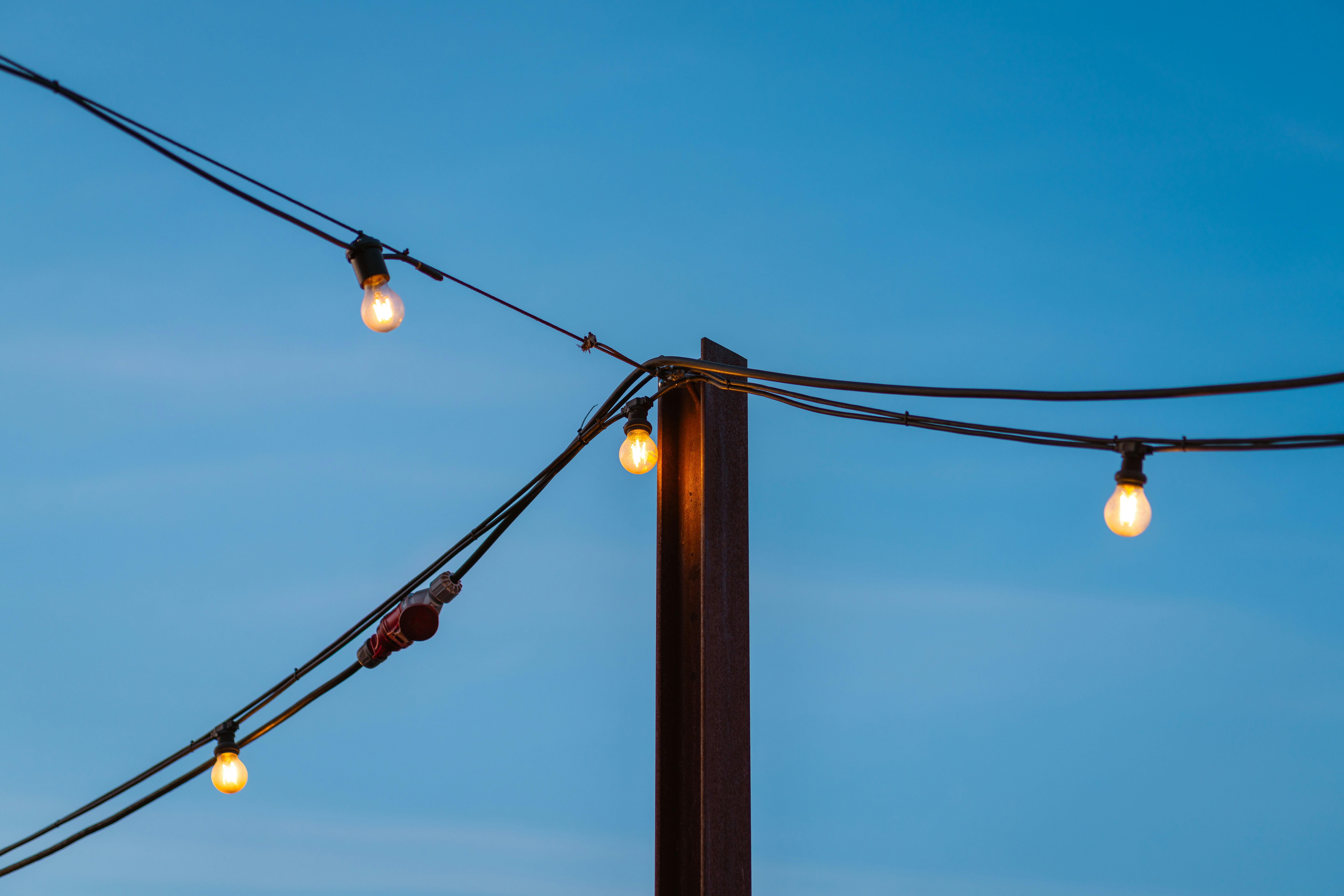 Several glowing bulbs strung along wires wrap around a wooden utility pole against a clear blue sky.