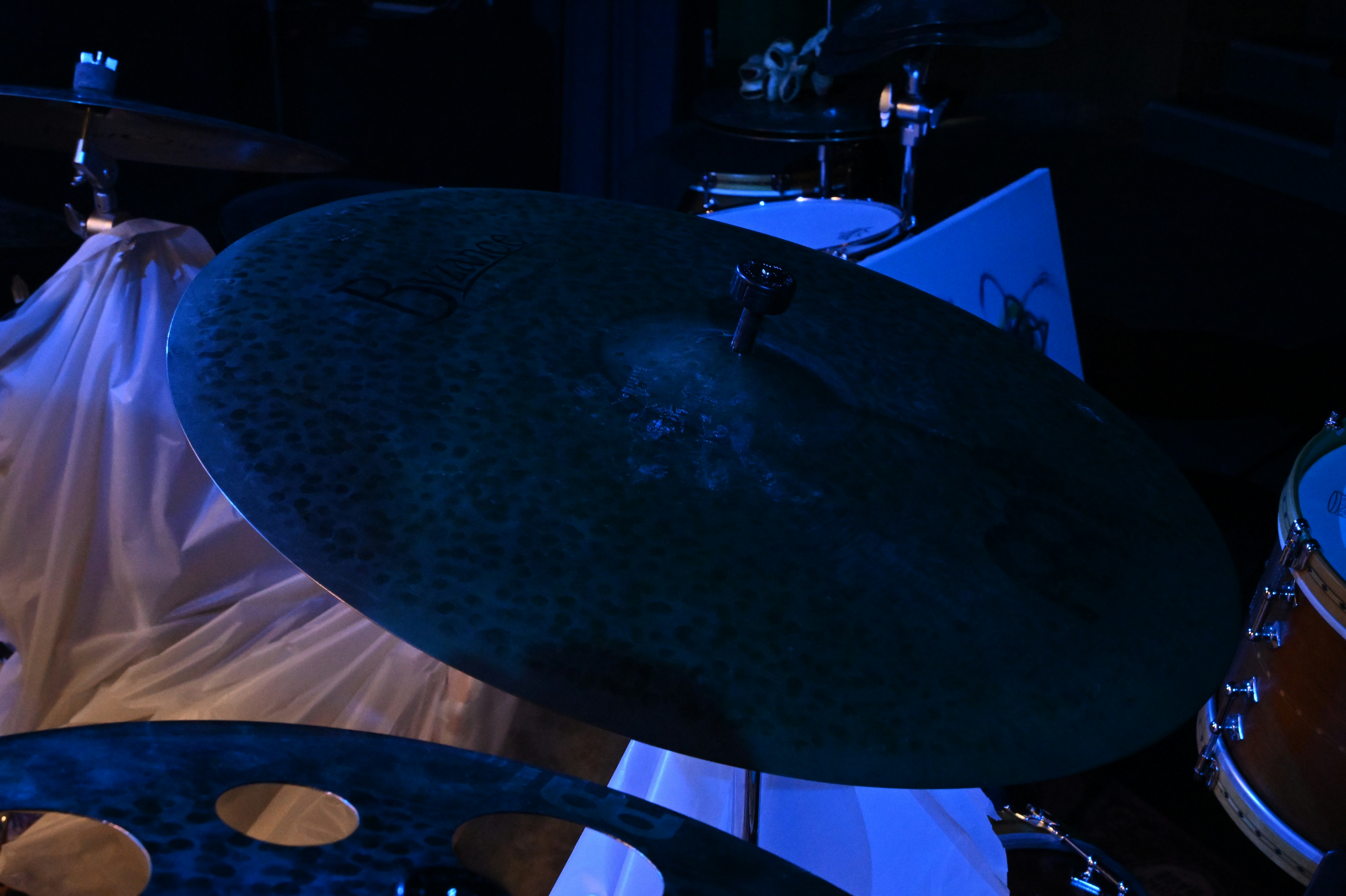 Blue-tinted drum kit on stage, cymbals catching ambient light in a low-lit concert setting. Clear focus on the drum array and towel-draped toms to convey a live performance mood.