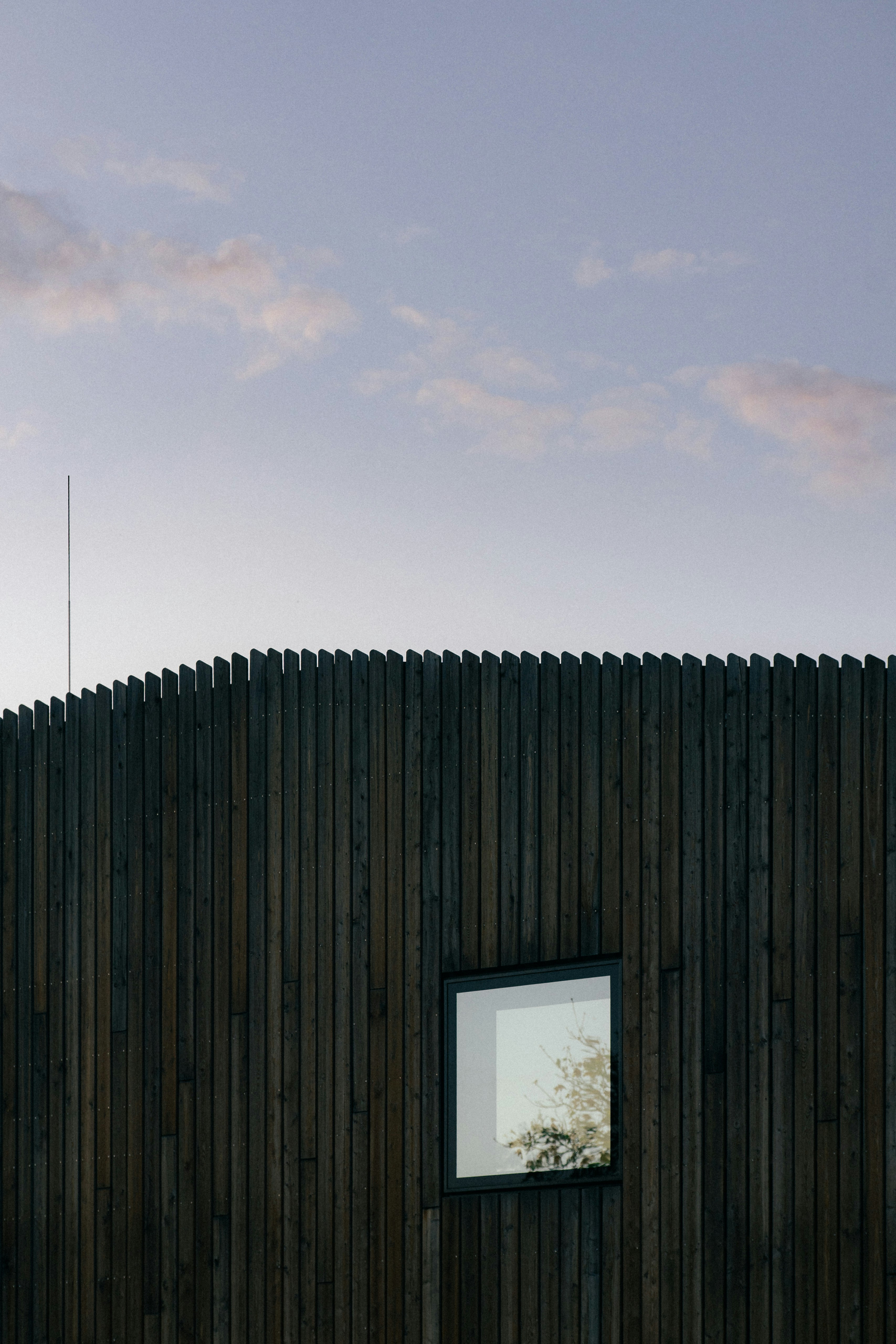 Dark wooden slatted wall with a single square window reflecting a hint of trees, under a pale sky. The image emphasizes architectural texture and minimal composition.