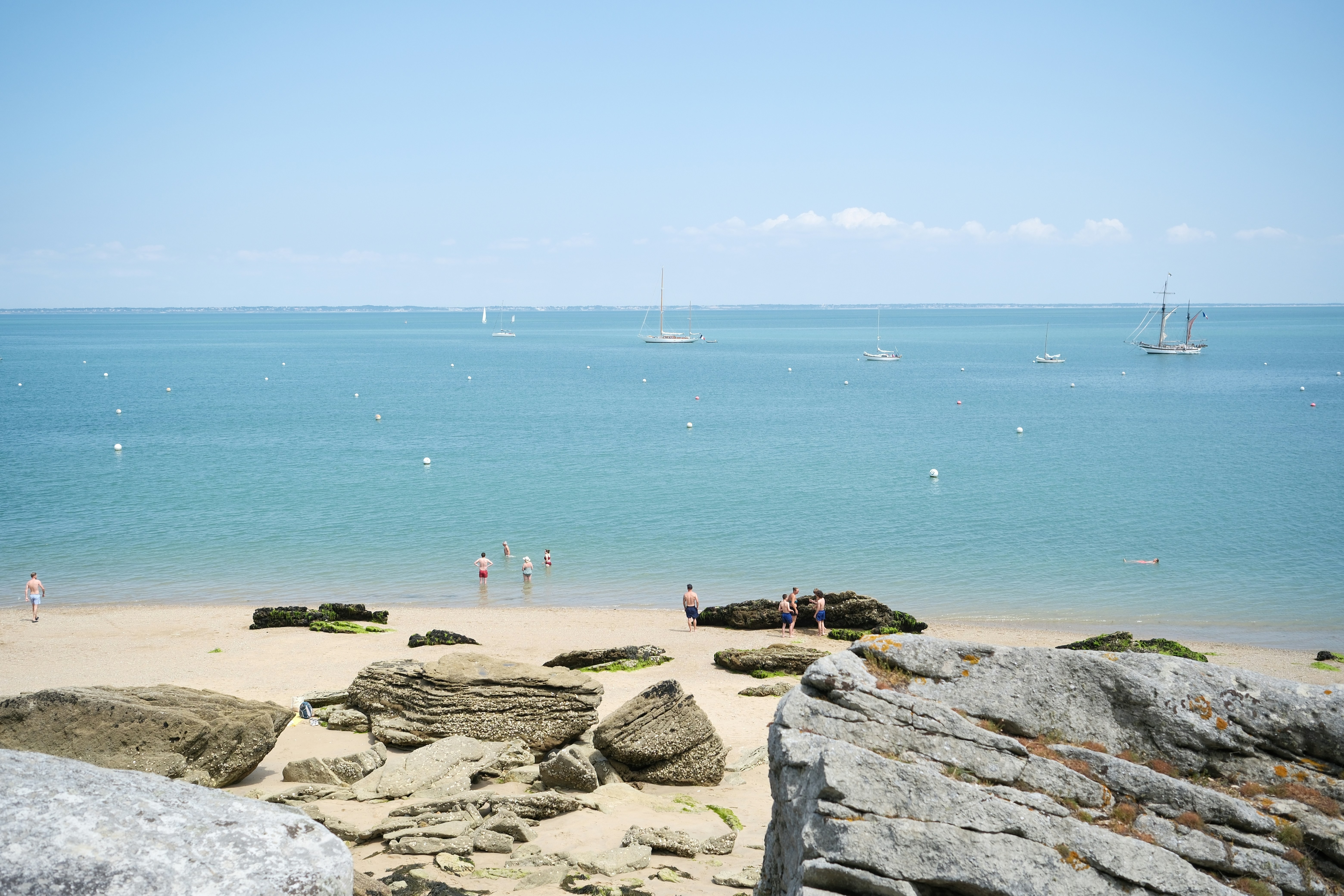 Quiet beach scene with swimmers near rocky shoreline and distant sailboats on calm turquoise water.