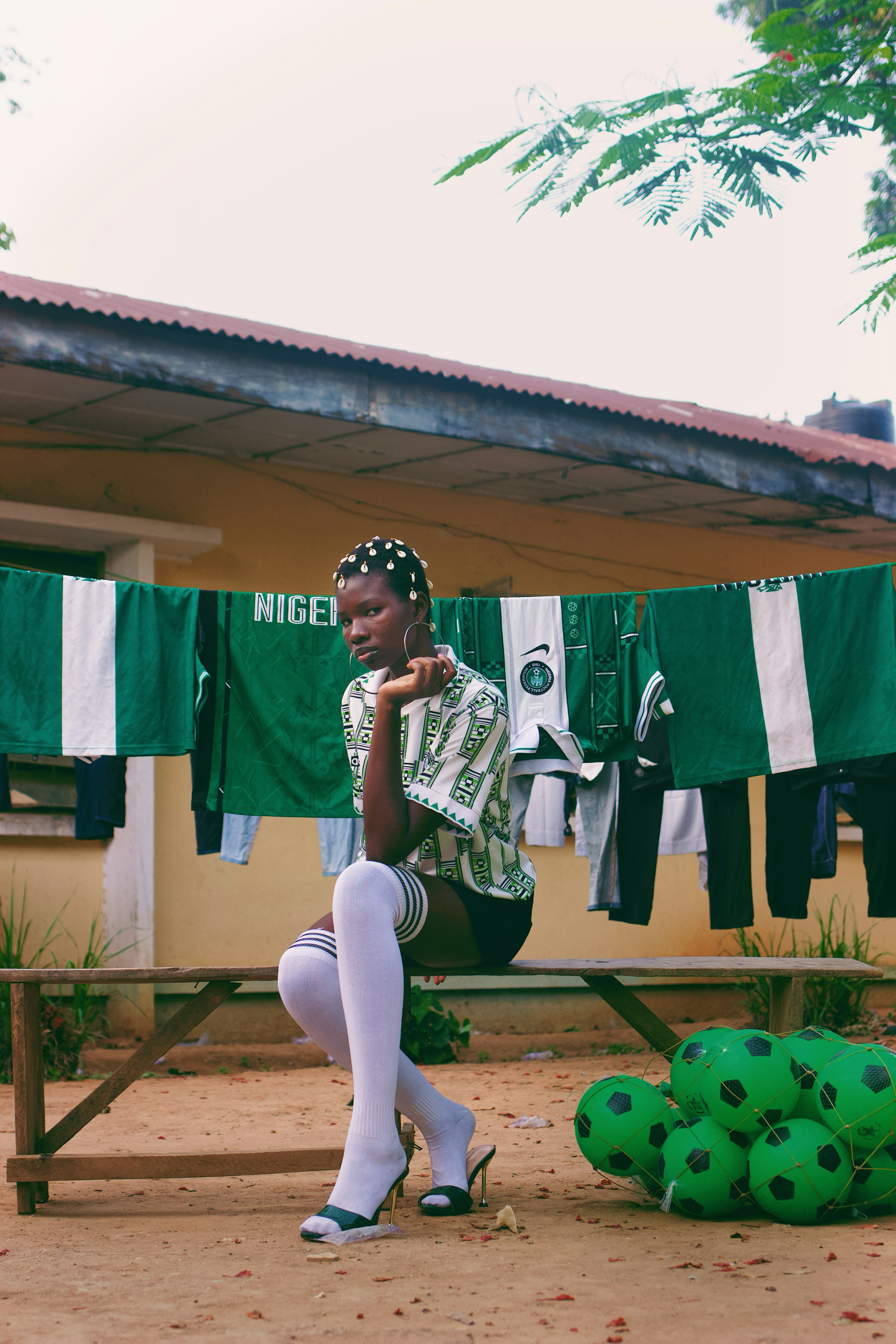 Young woman seated in front of green-and-white laundry clothesline, with green soccer balls on sand nearby; outdoor scene with a yellow building backdrop.