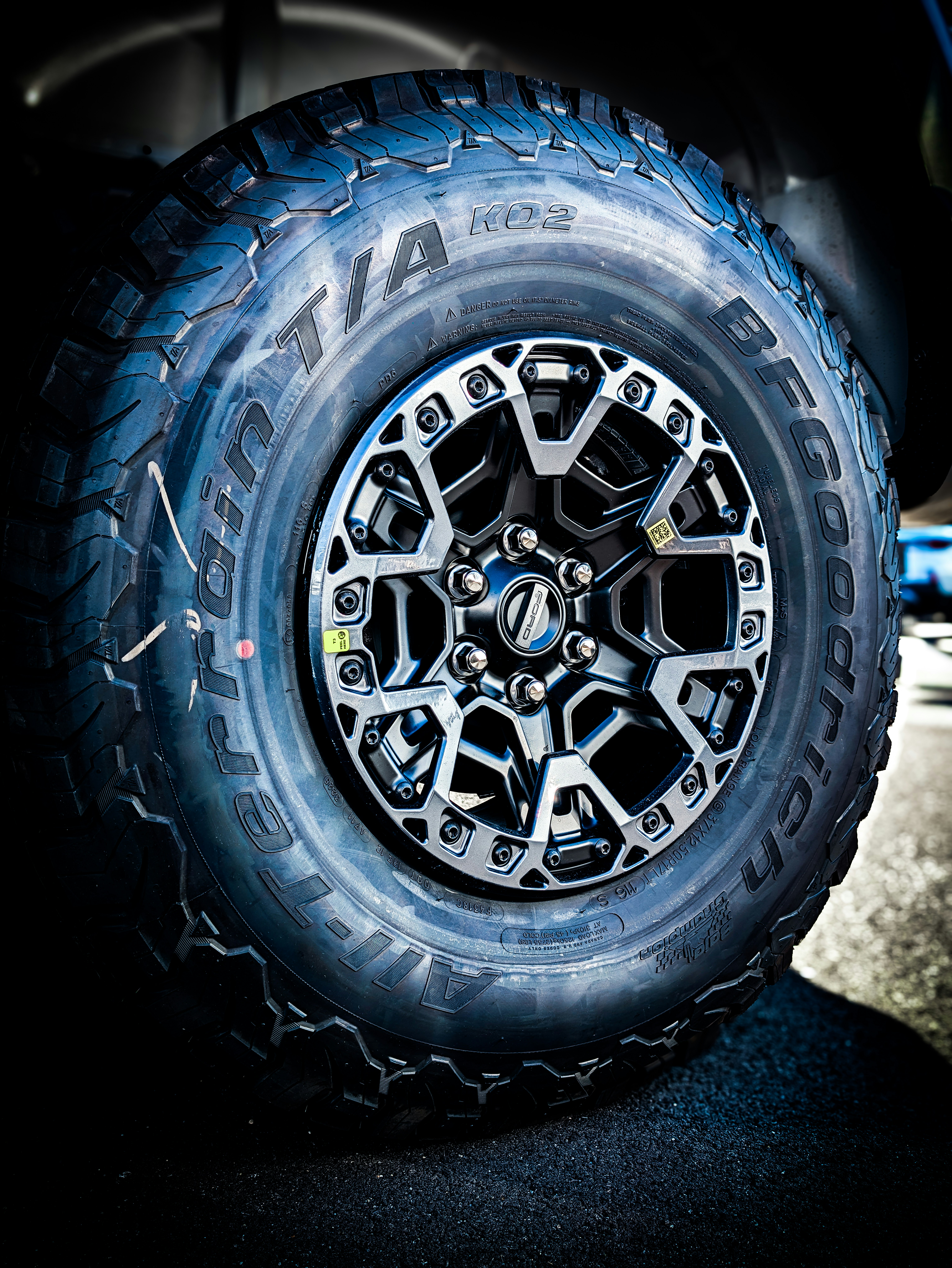 This macro photograph details a rugged off-road tire featuring deep tread blocks, securely mounted on an intricate two-tone alloy wheel, illuminated by crisp highlights against a heavily shadowed automotive background.