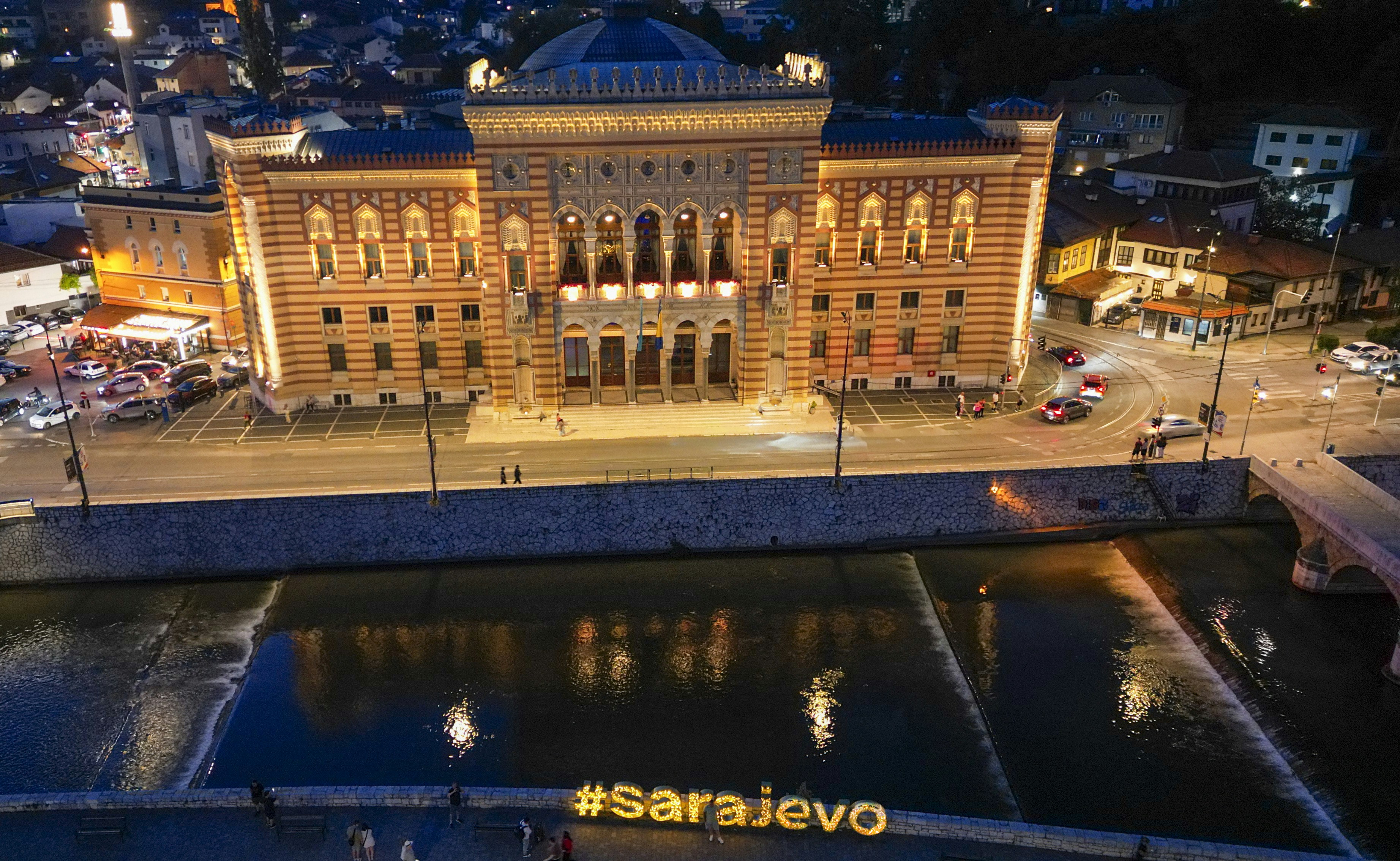 Aerial view of Sarajevo’s grand city hall illuminated at dusk, with the river and reflective water foreground and the hashtag sculpture '#sarajevo' along the riverbank.