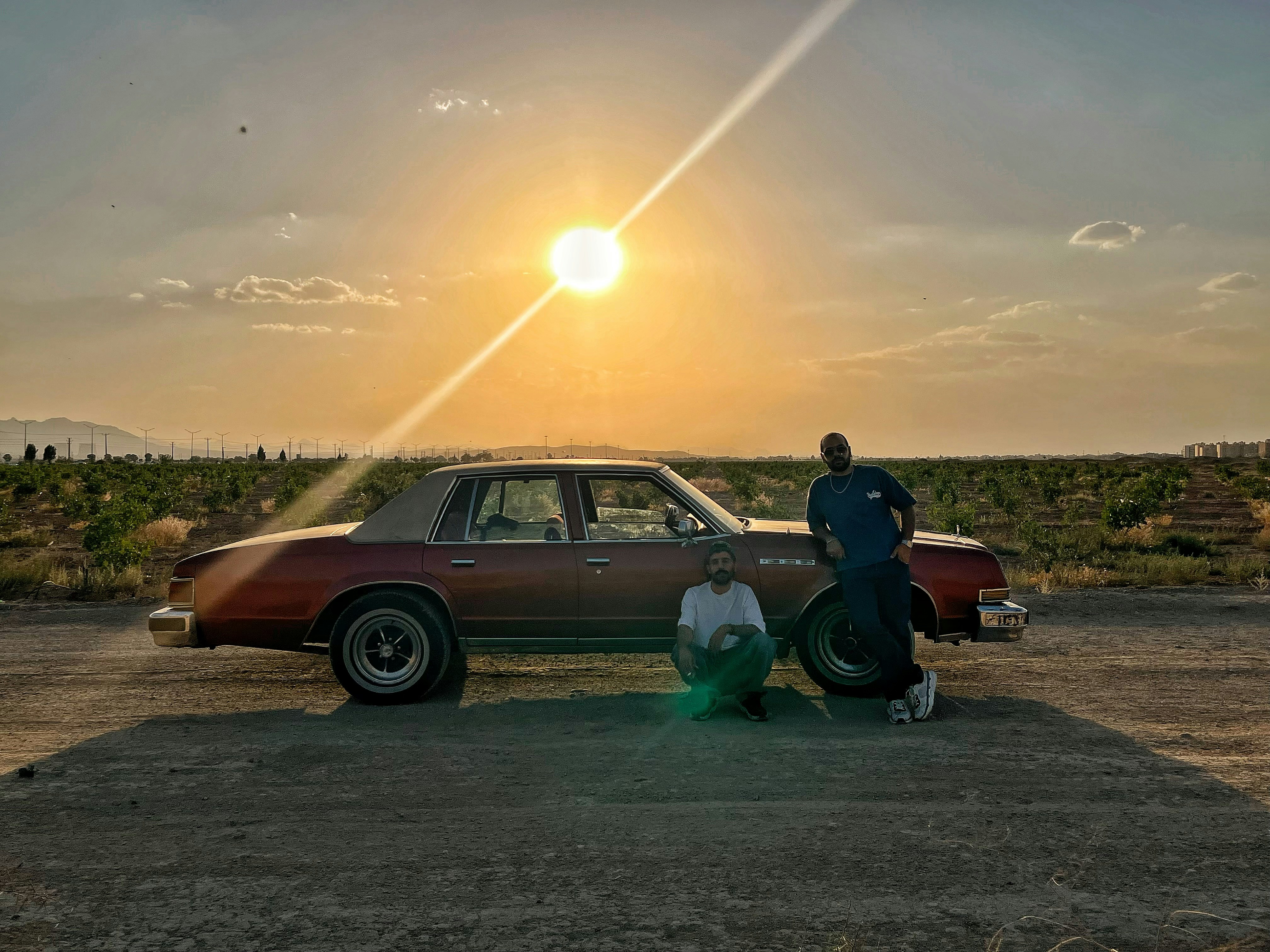 Two men pose beside a vintage car under a low desert sun, with a long horizon and a warm glow emphasizing the scene. A logo-like emblem on the car hints at a vintage vibe.