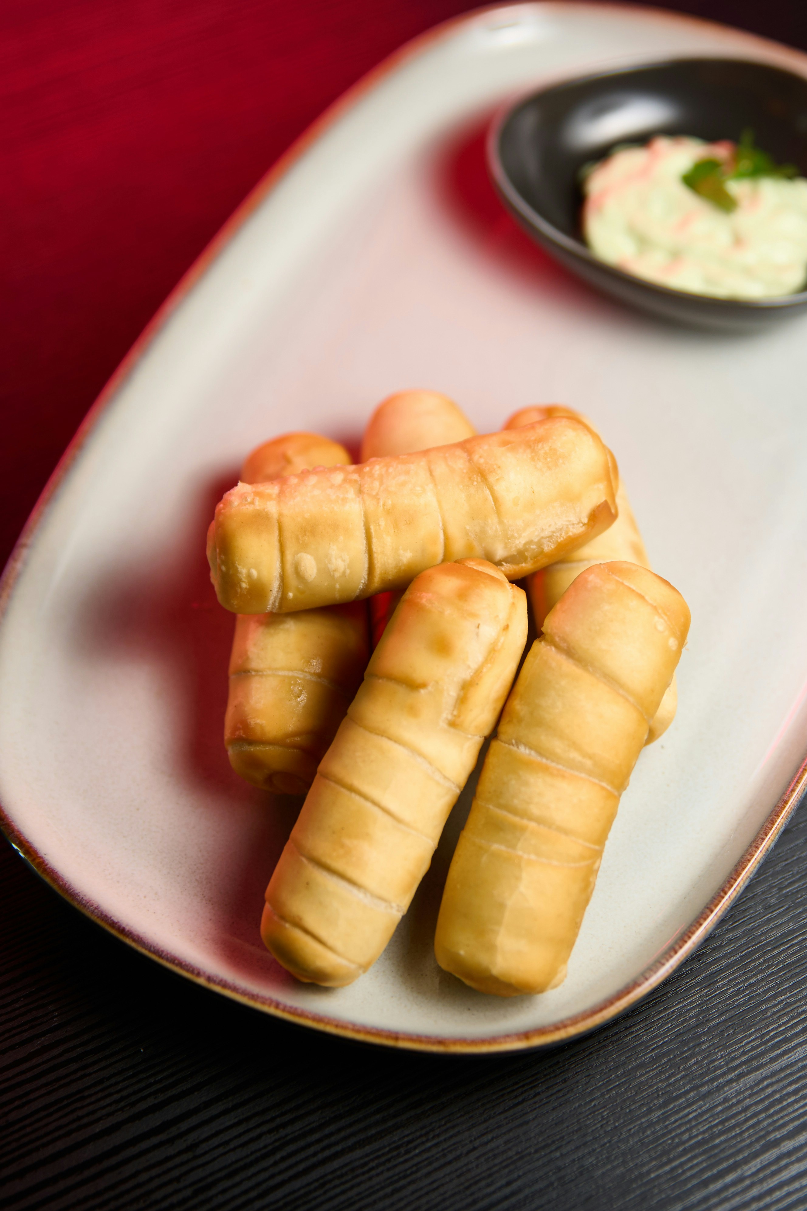 Golden-brown finger rolls arranged on a pale plate with a small bowl of dipping sauce in the background. Food-focused composition highlighting texture and color, presented as a plated snack.