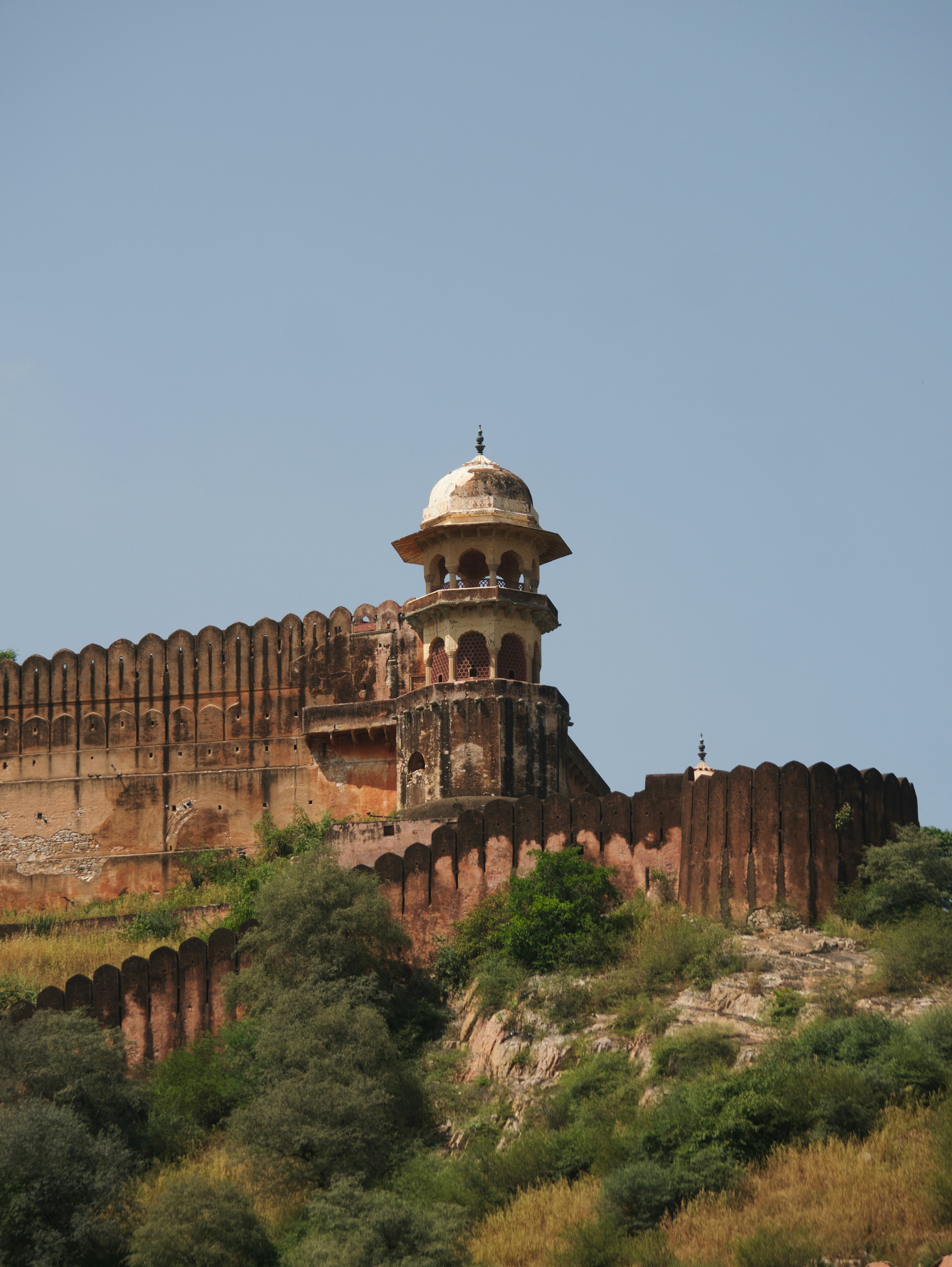 A historic fortress on a hillside with weathered walls and a domed turret, rising above greenery under a clear blue sky.