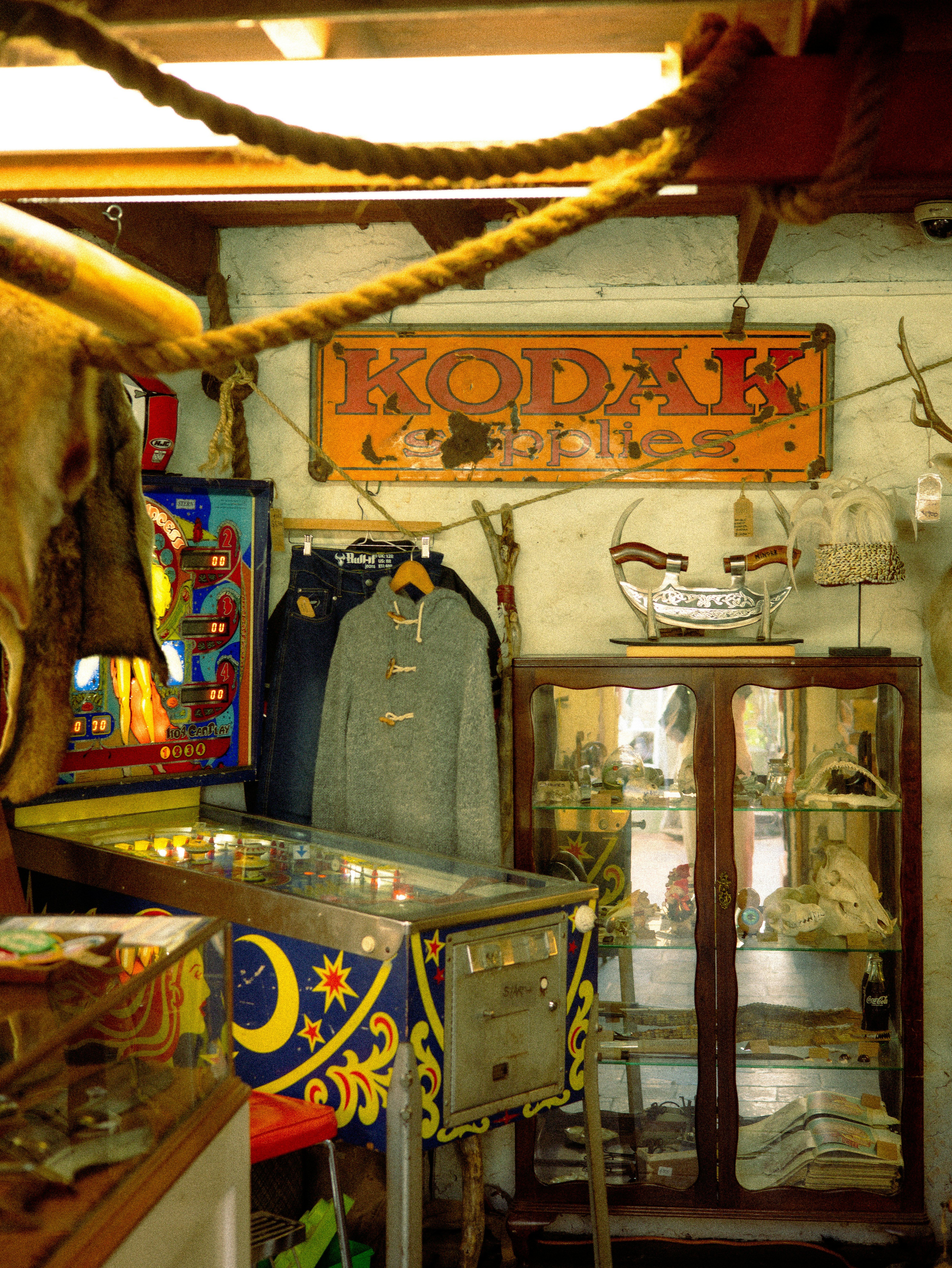 An eclectic vintage shop interior featuring a weathered Kodak sign, a glass-display cabinet, and retro arcade elements. A gray duffle coat hangs center, with old toys and bright patterned furnishings surrounding a glass-topped counter.