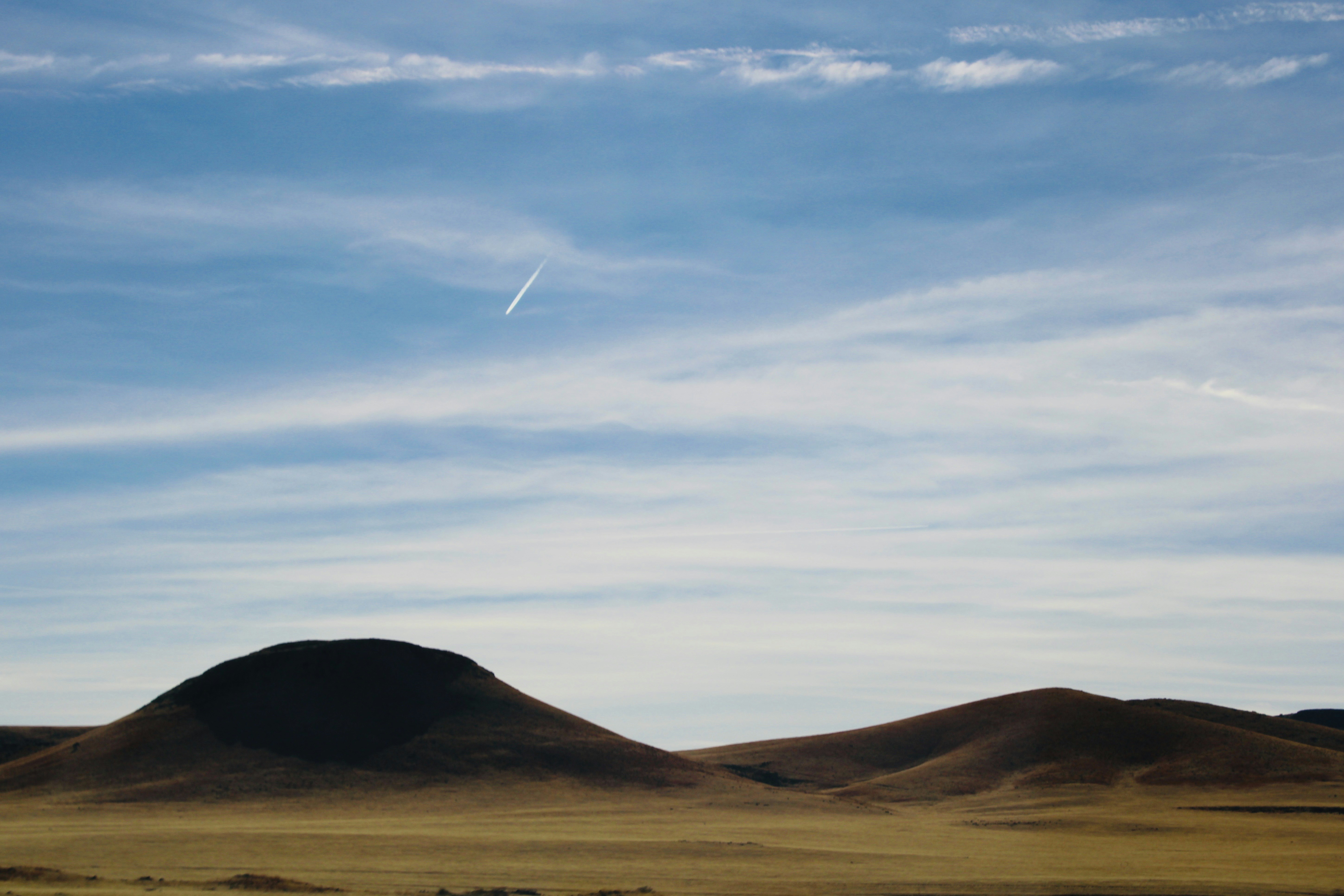 Low wide-angle landscape of rolling desert hills under a layered blue sky with wispy clouds and a faint contrail; the scene emphasizes wide open space and subtle color gradients.
