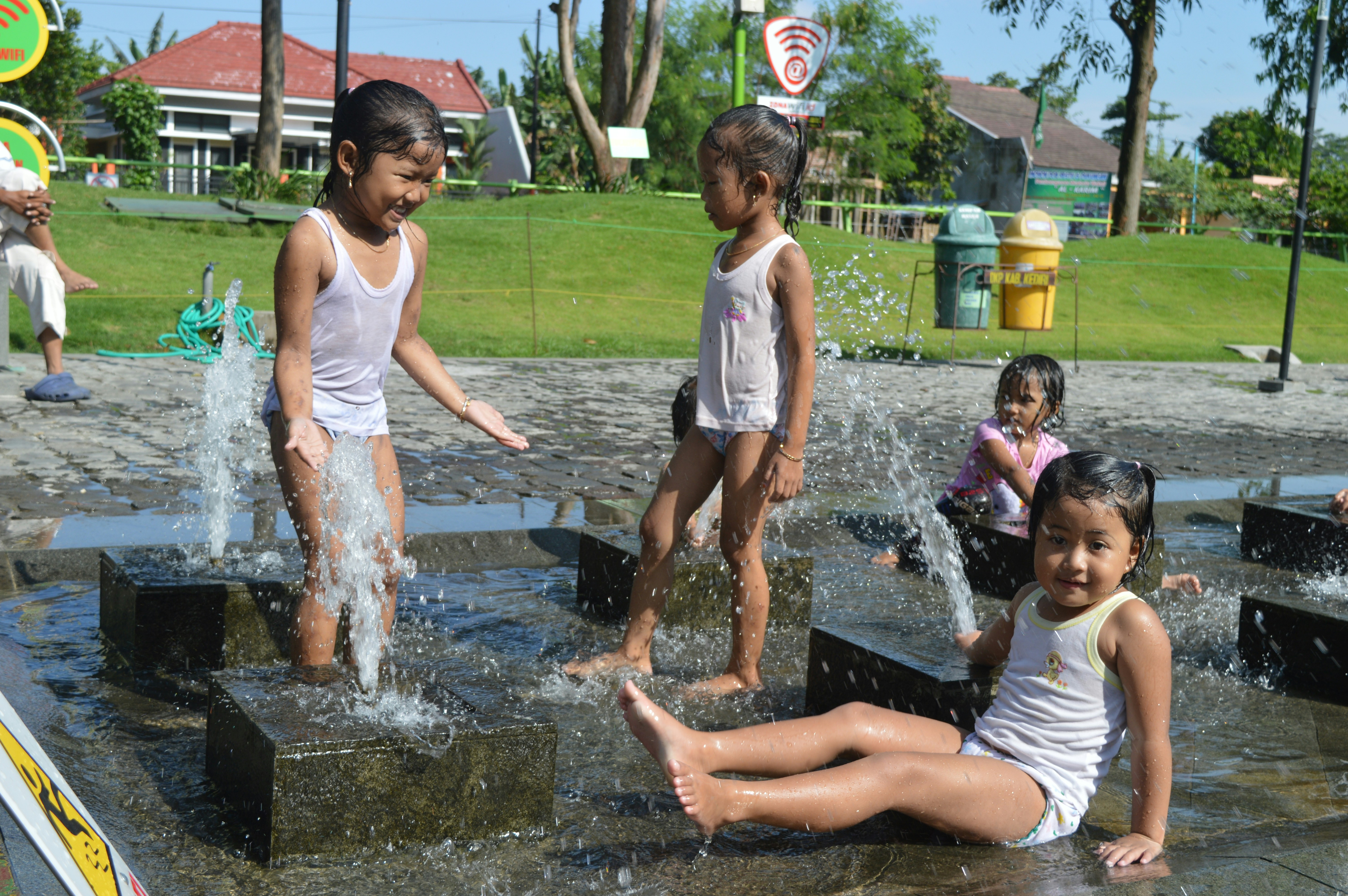 Group of kids in white outfits playing in a shallow public fountain with water jets, on a sunny day in a park-like setting; a seated girl in the foreground and others mid-play near the water features.