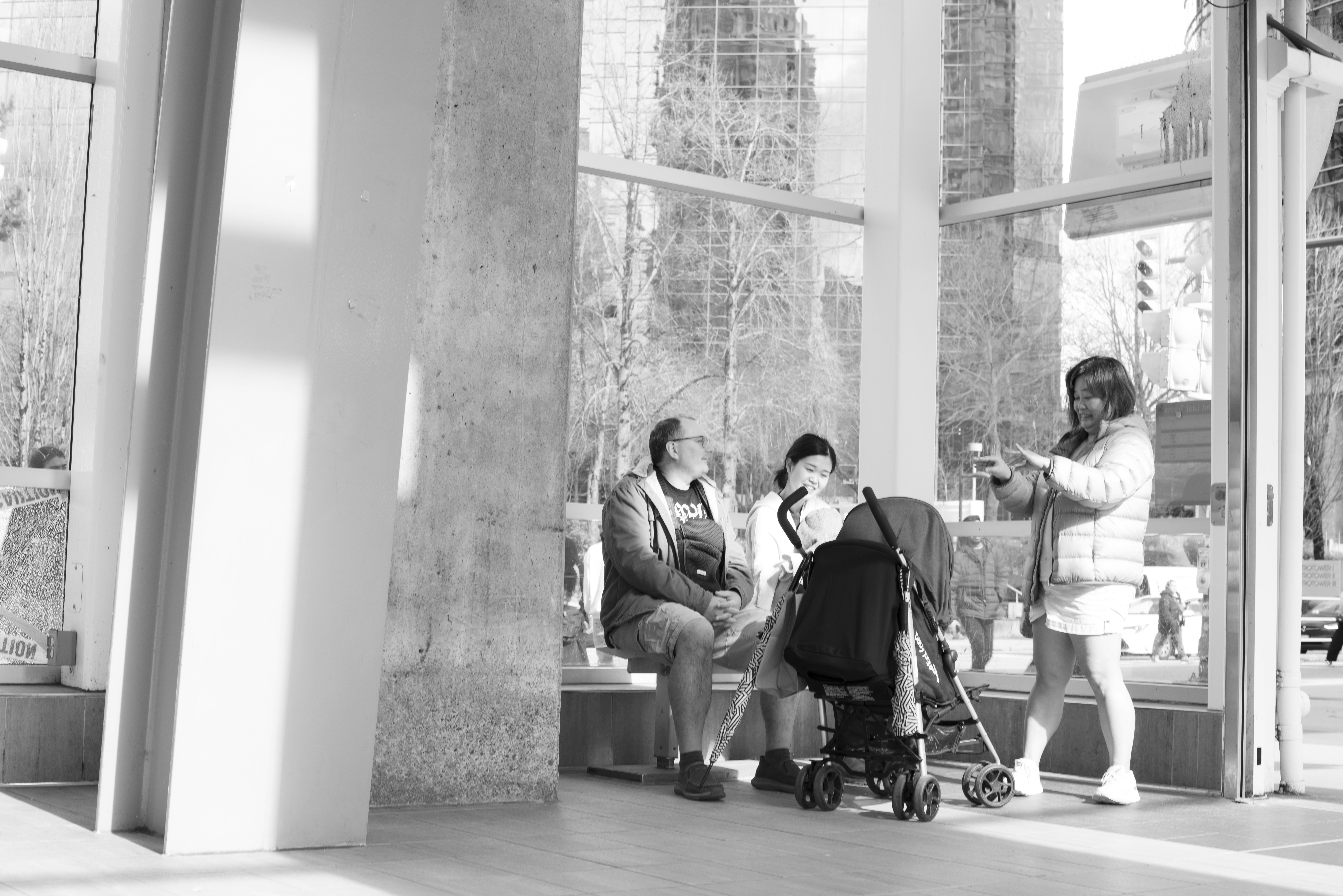 People sit and chat near a stroller inside a bright, glass-walled atrium; a person on the right appears to be taking a photo. A stroller and minimal furnishings anchor the scene.