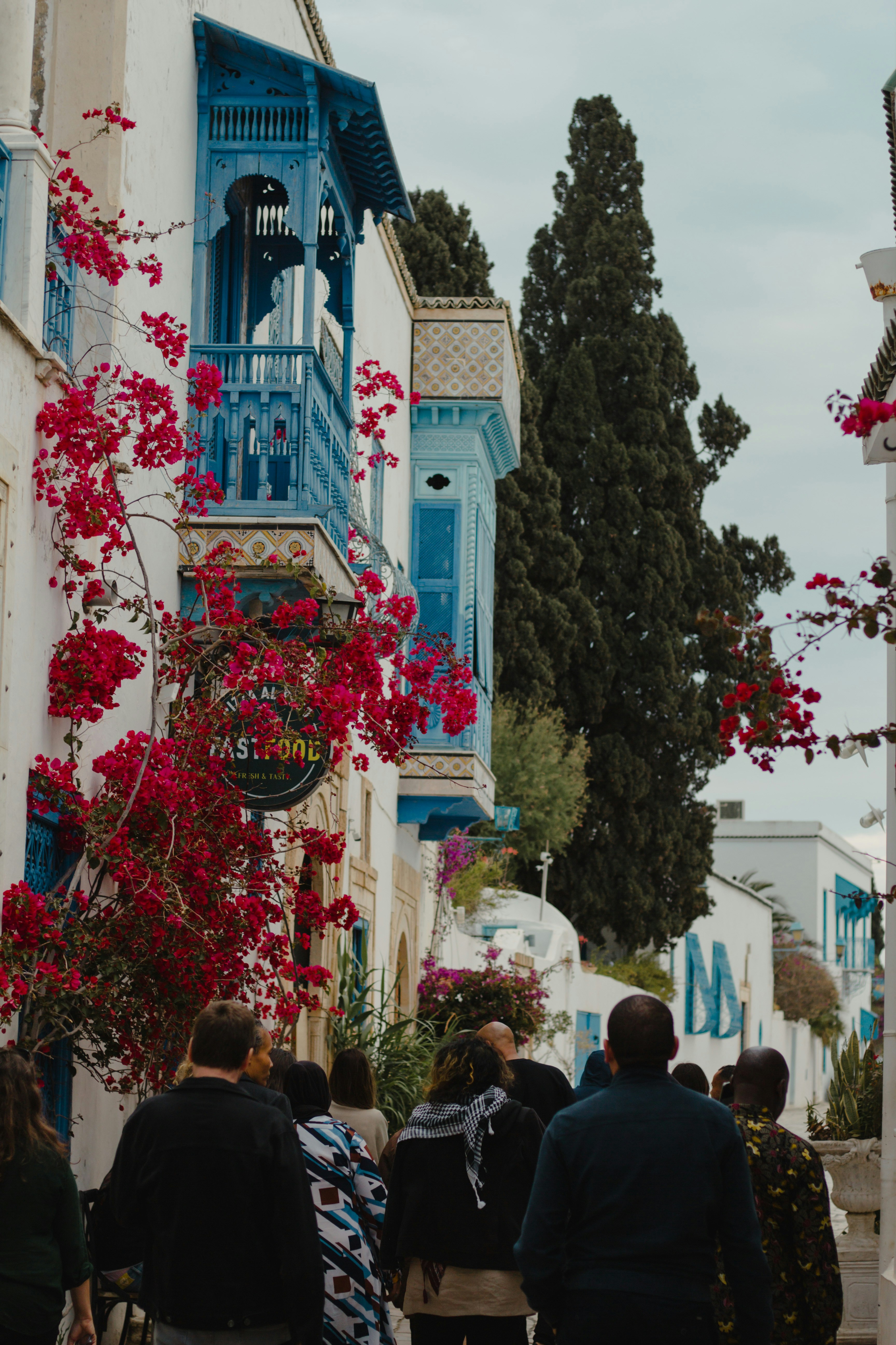 Colorful blue balconies frame a sunlit street with a crowd of people below; vibrant bougainvillea climbs the white walls, yielding a lively urban scene.