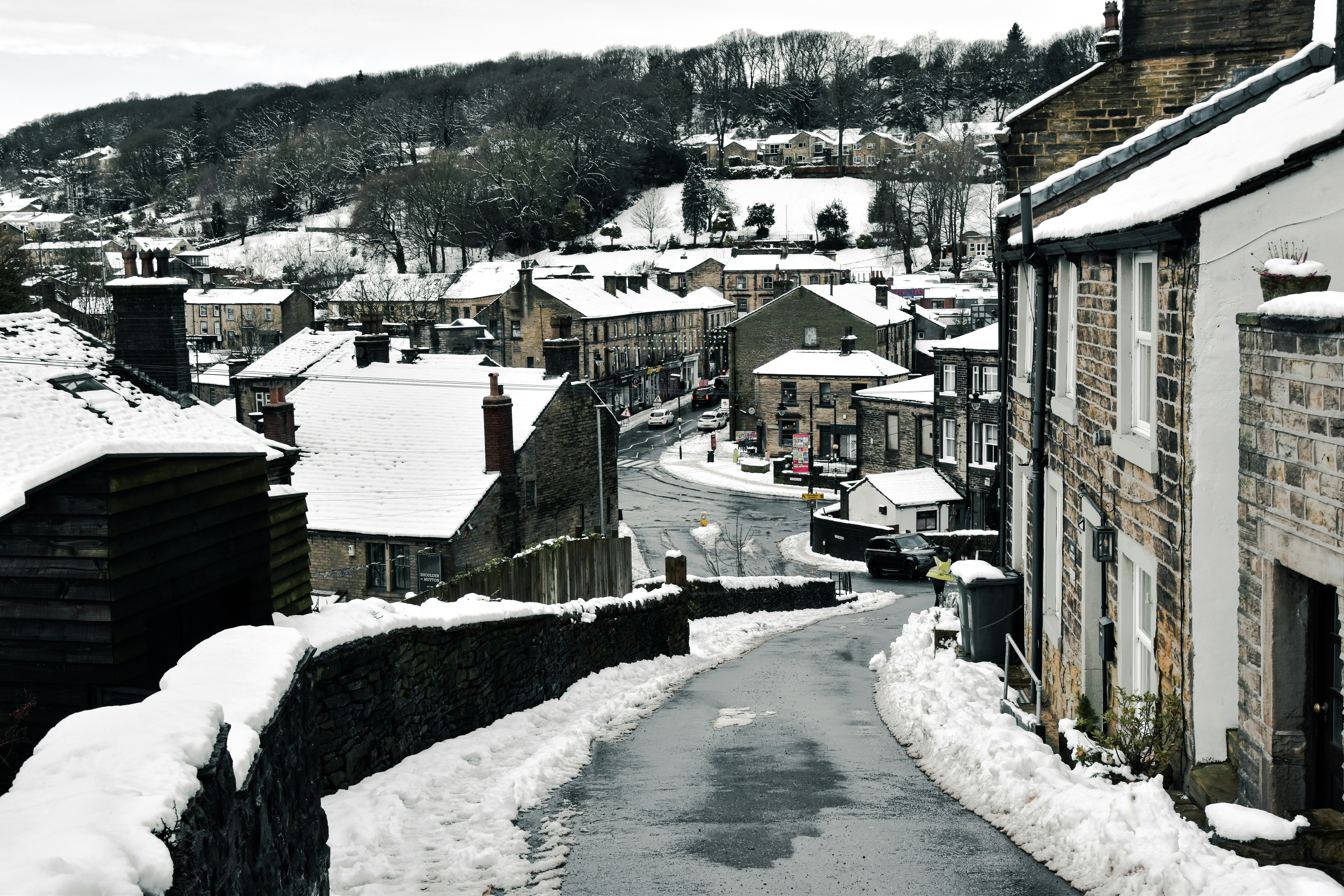 A scenic high-angle view looking down a narrow, snow-lined sloped street towards Holmfirth town centre in West Yorkshire after a fresh snowfall. Fresh snow blankets the roofs of traditional stone cottages and surrounding hillside, capturing a serene winter scene across the historic Pennine valley town.
