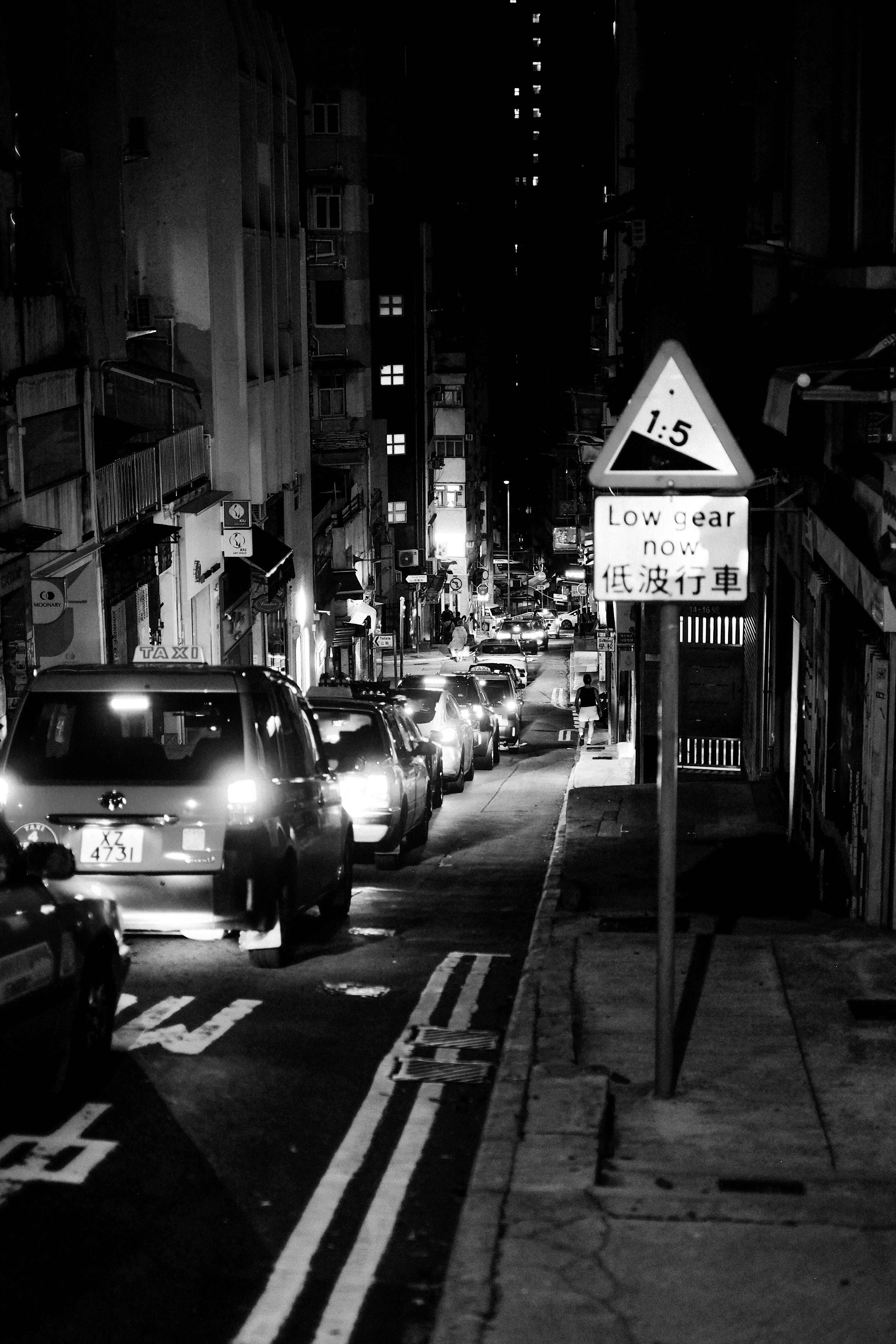 Monochrome city street at night with cars lined along a narrow avenue and a prominent signpost in the foreground.