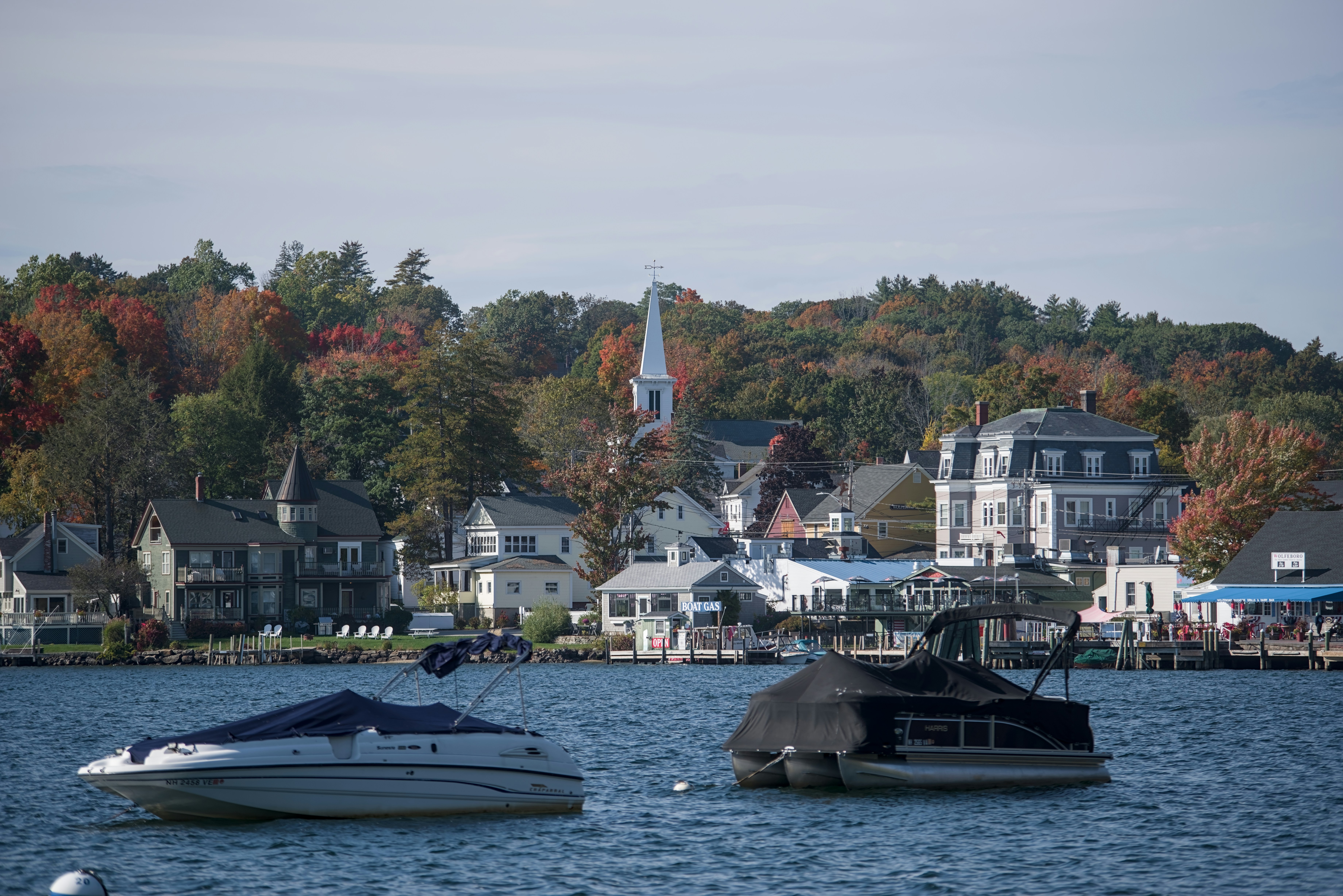 Boats float on calm water with a harbor village and autumn trees in the background, conveying a quiet lakeside scene.