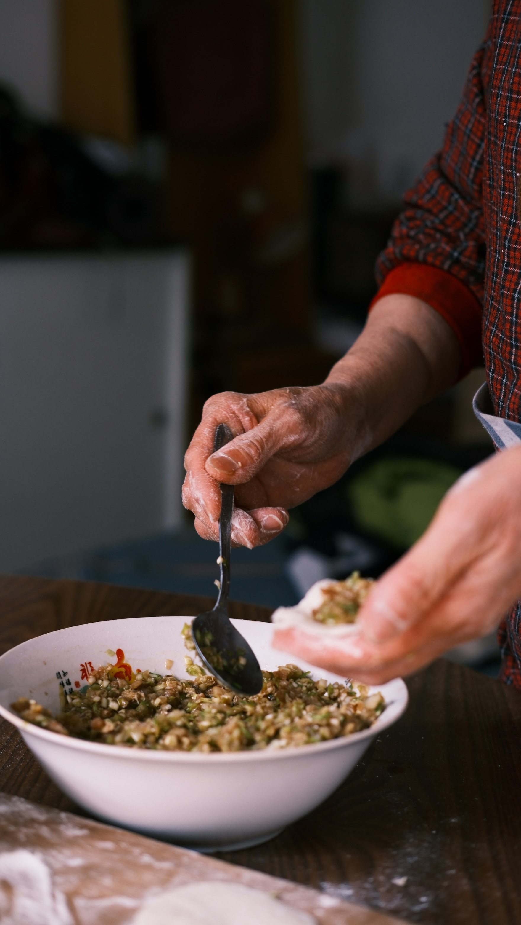 A close-up of hands preparing a bowl of chopped filling at a kitchen counter, with flour-dusted surface and a wooden table in warm indoor light. A logo-like pattern on the bowl hints at cultural cuisine.