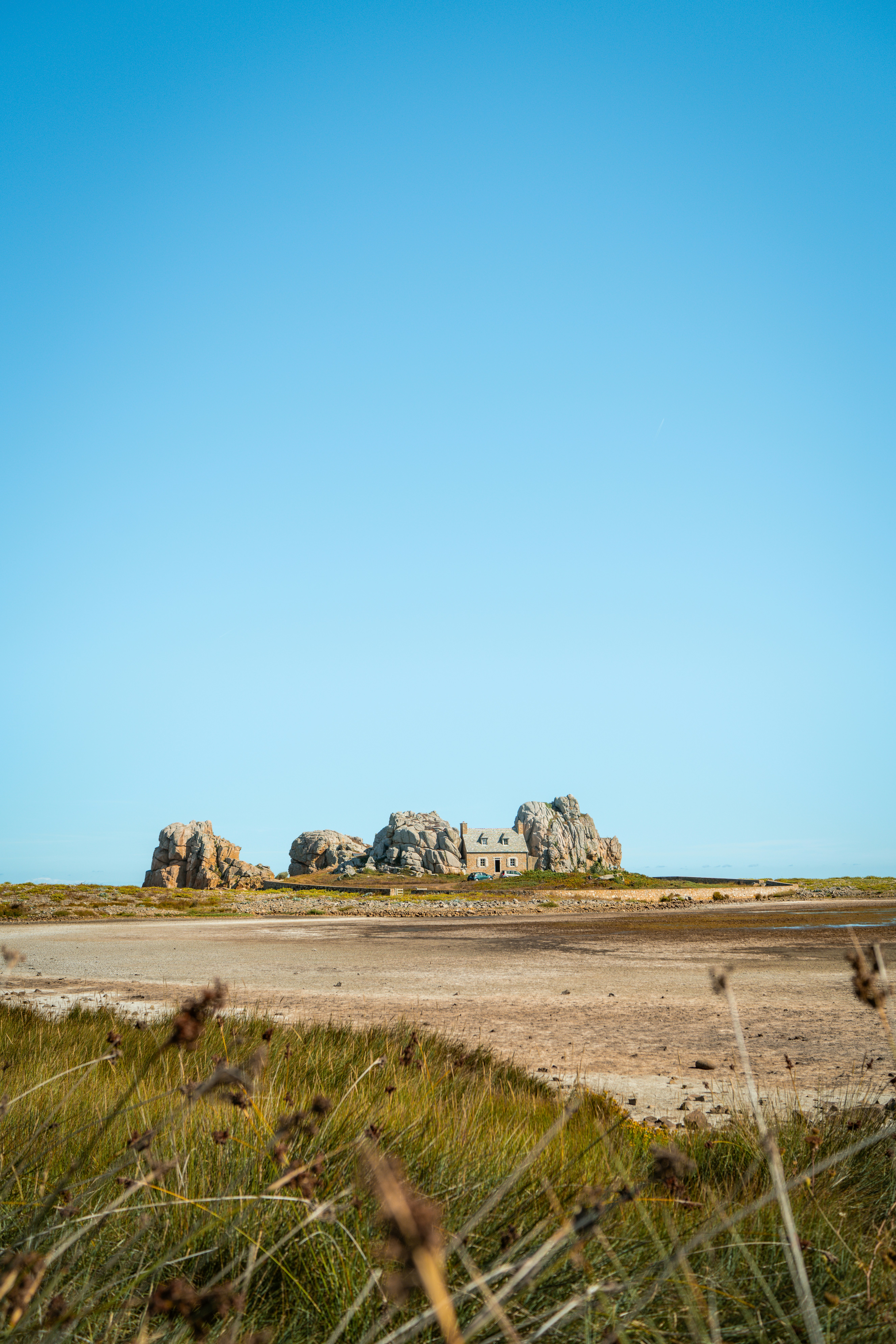Coastal landscape with a small stone house framed by rocky outcrops on a grassy shore under a bright blue sky. A wide, serene seascape emphasizing the distant structure and natural foreground.
