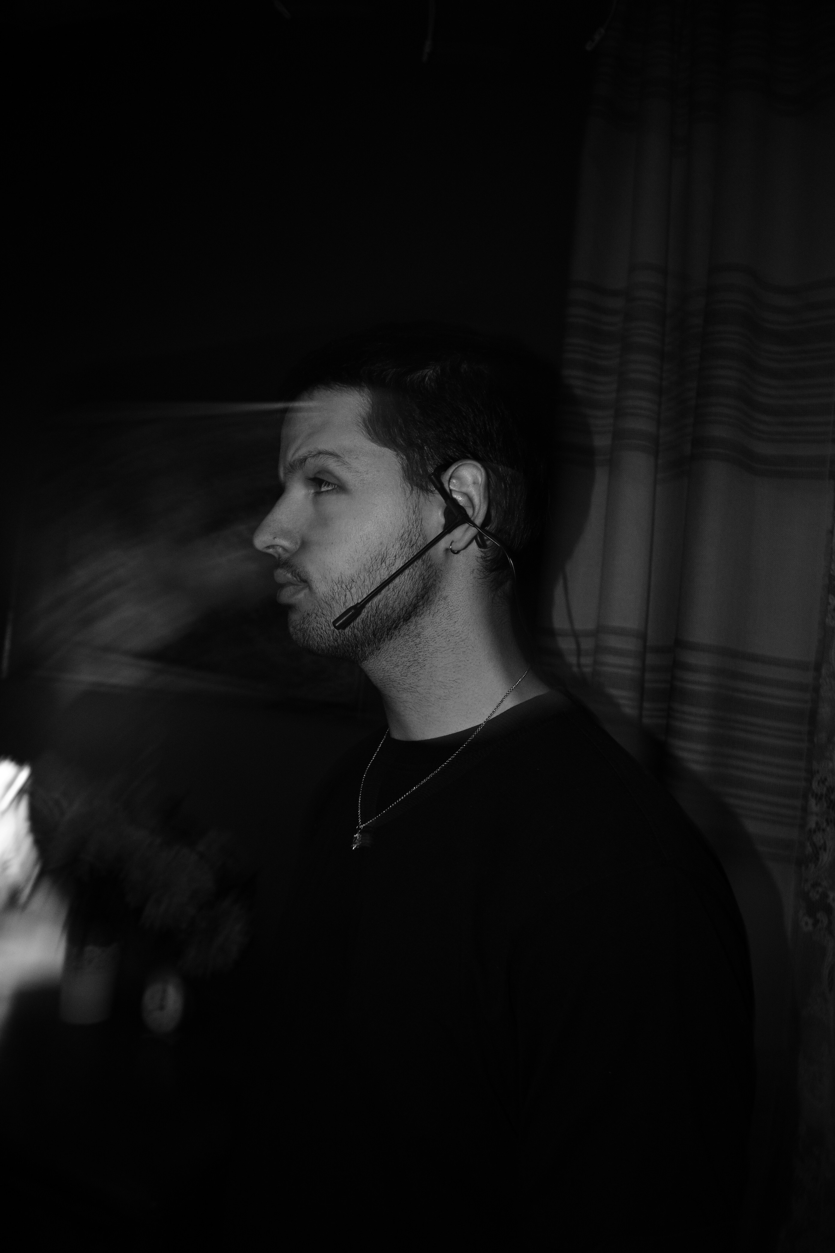 A man wearing a headset microphone stands in low light, head turned to the side with motion blur suggesting movement; curtain backdrop and faint foreground light contribute to a moody, documentary vibe.