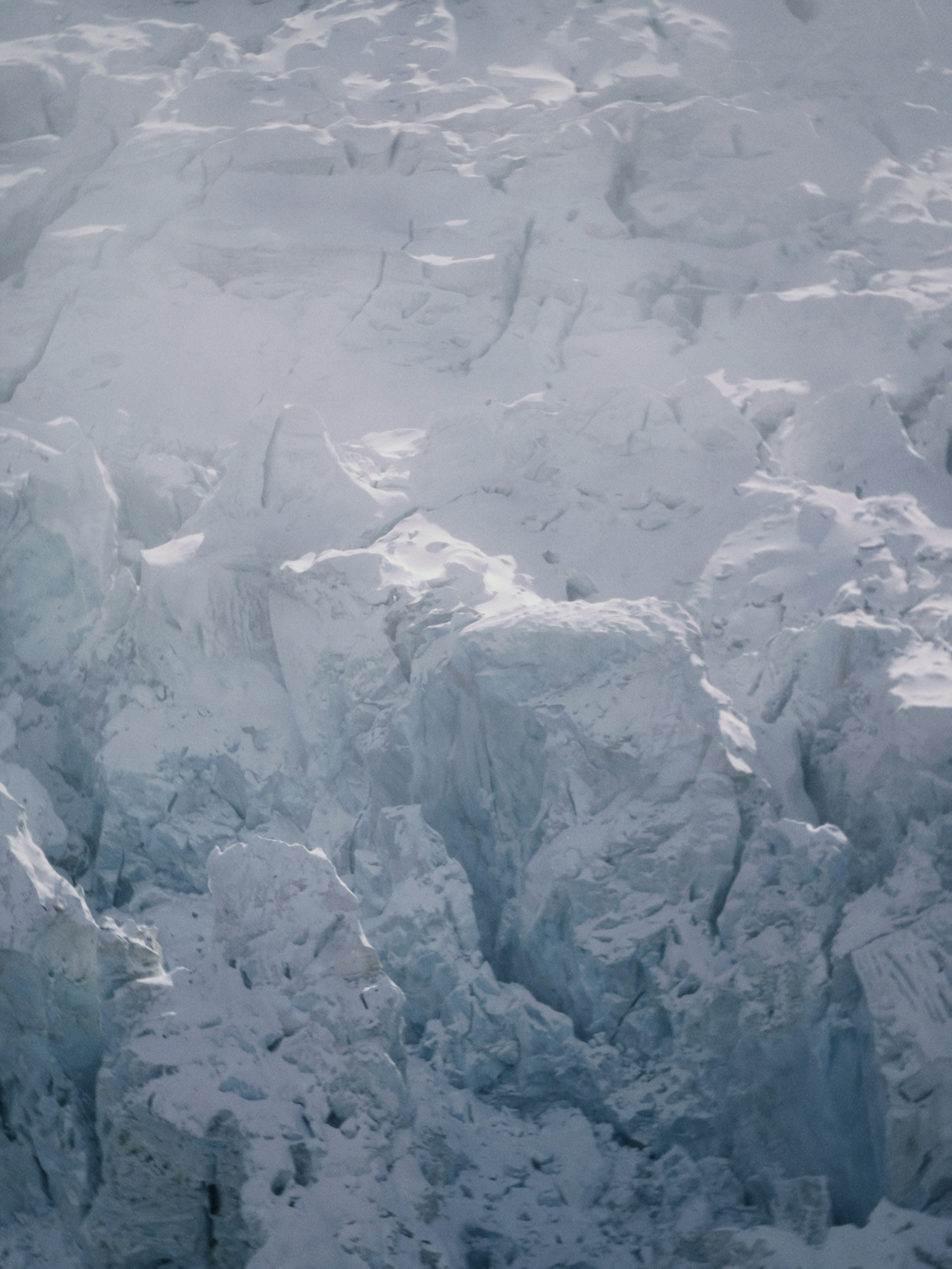 Glacial terrain with jagged ice formations and soft blue shadows; a stark, sculpted ice face suggesting a rugged glacier landscape.