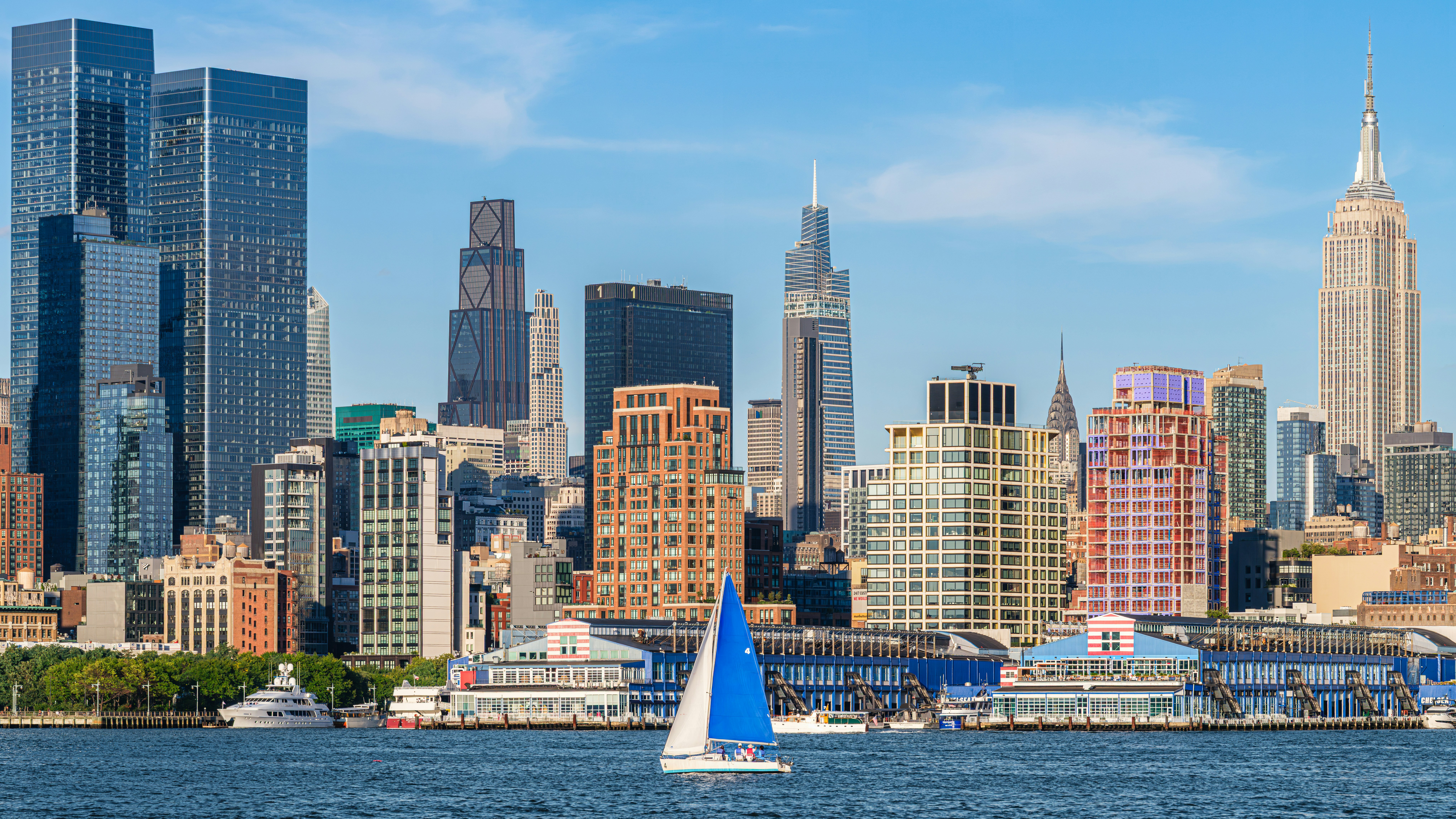 Sailboat with blue sail glides near a dense city skyline along the waterline; foreground features calm harbor and a pier, with iconic skyscrapers in the background.