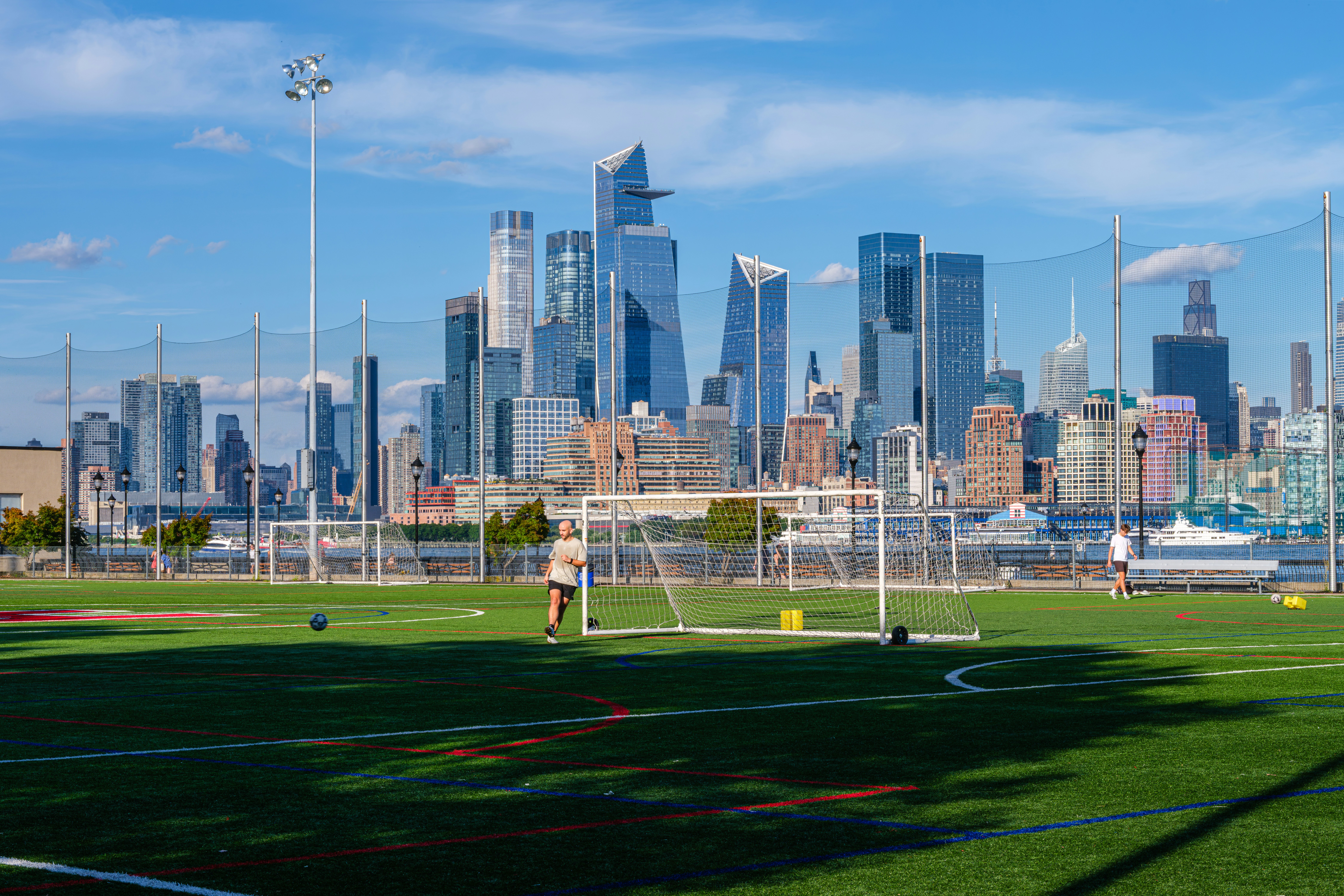 Soccer field with players and goalposts set against a modern city skyline skyline behind, under a blue sky.